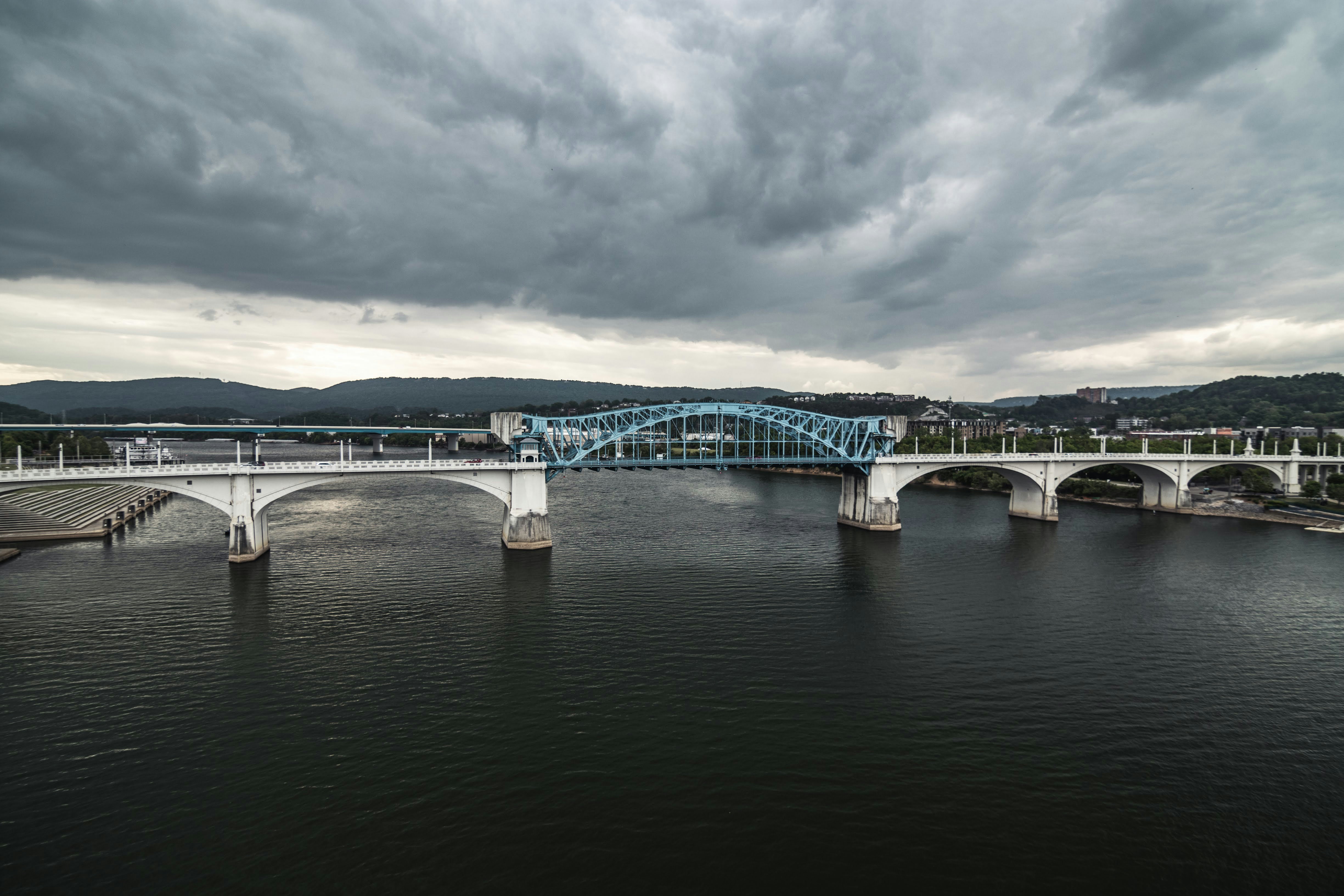 A striking blue bridge arches over a calm river, framed by dark clouds and distant hills, showcasing urban architecture against a dramatic sky.