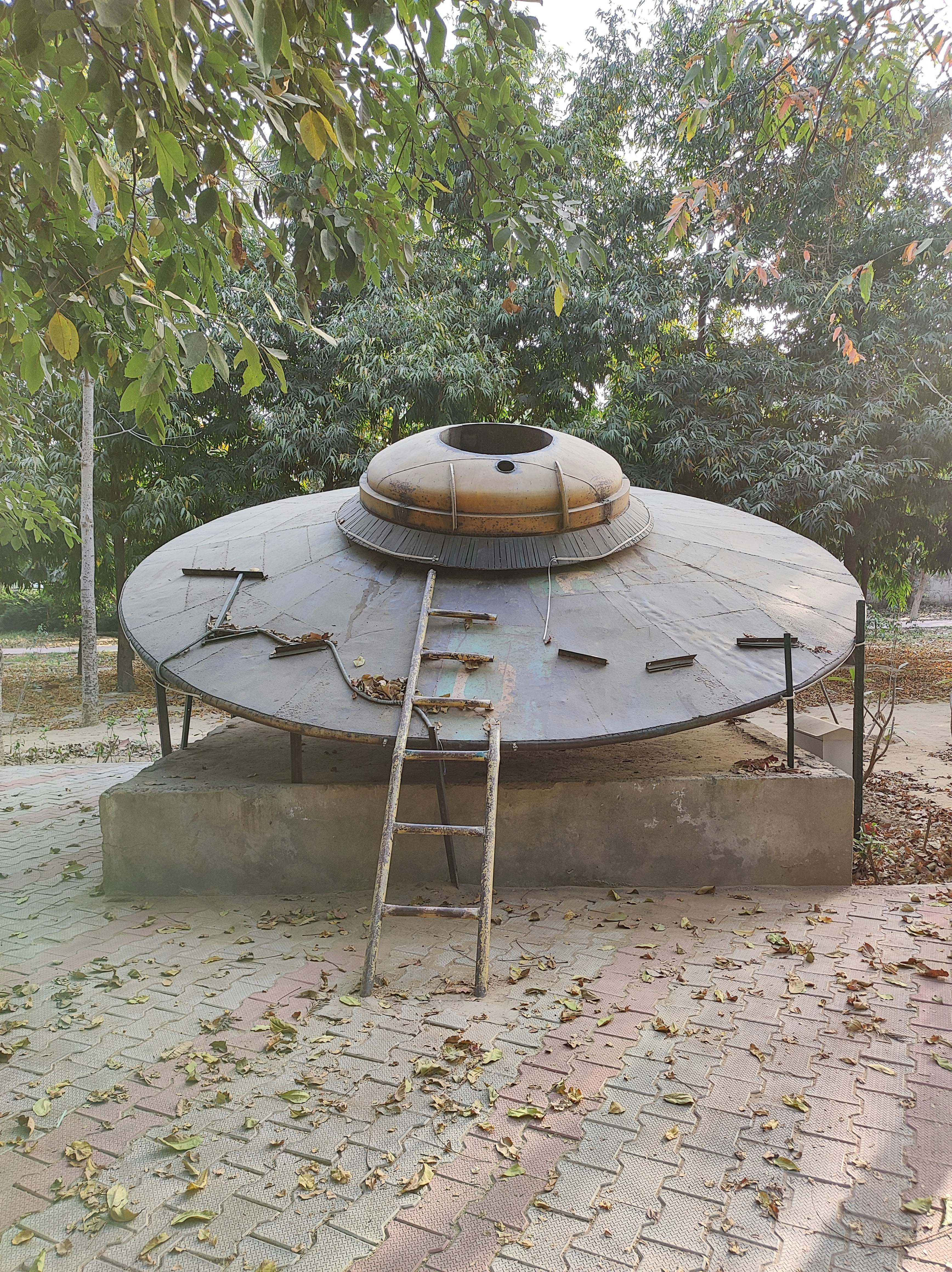 Weathered, disc-shaped metal structure sits on a concrete pedestal in a park, with a ladder leaning against it and fallen leaves scattered around.