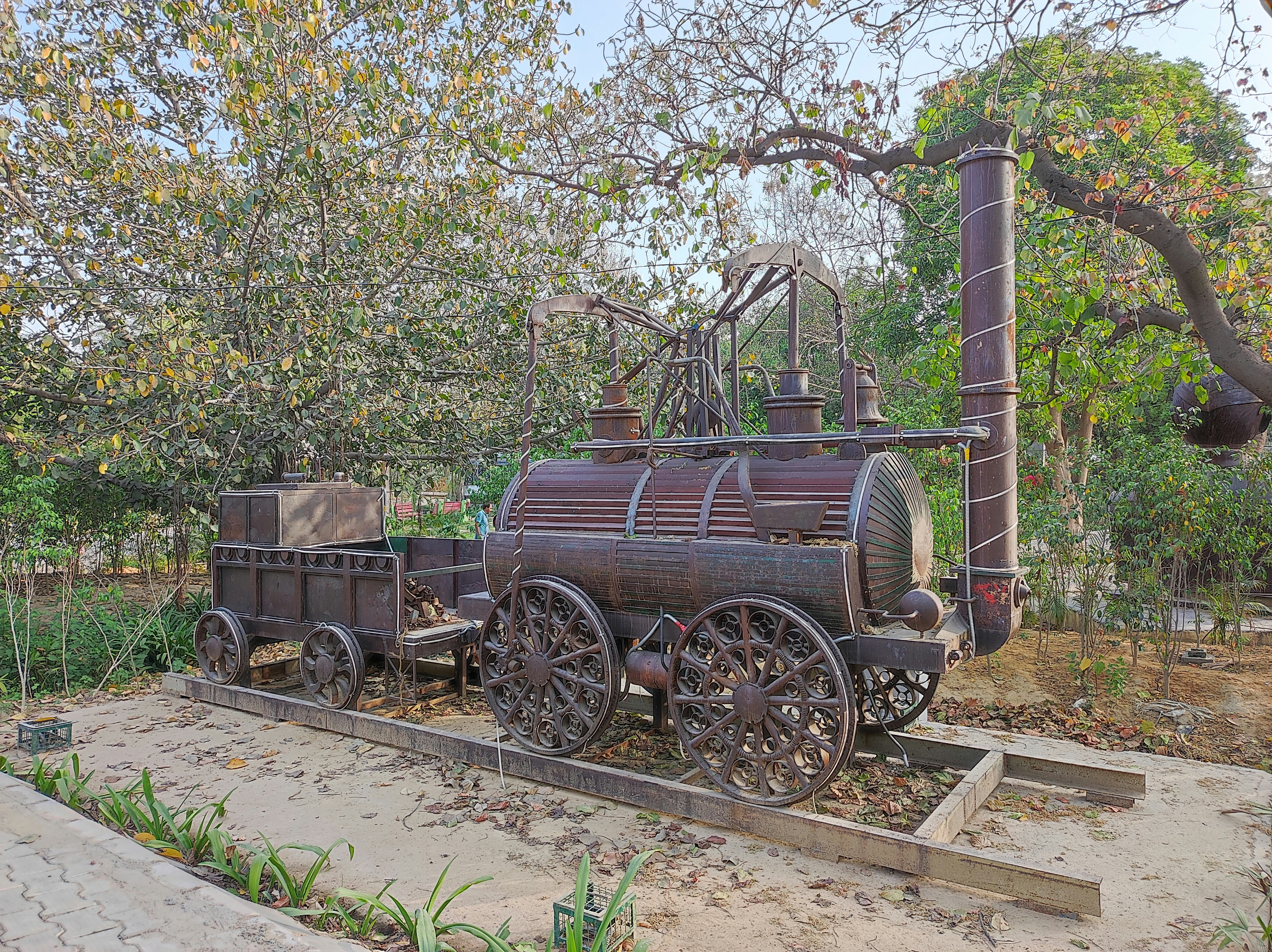 Old steam locomotive sculpture sits on a low platform in a park. The rusty engine is framed by trees and greenery, highlighting industrial heritage.