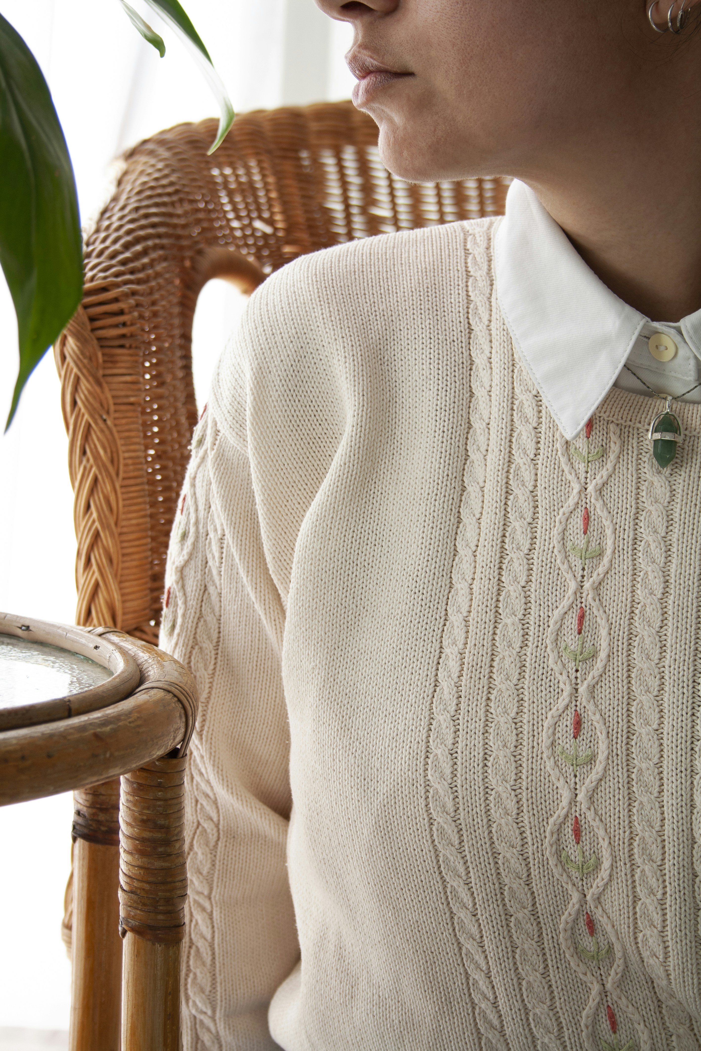 Close-up of a person wearing a knitted sweater and a collared shirt, seated beside a potted plant and a wicker chair, capturing a serene indoor atmosphere.