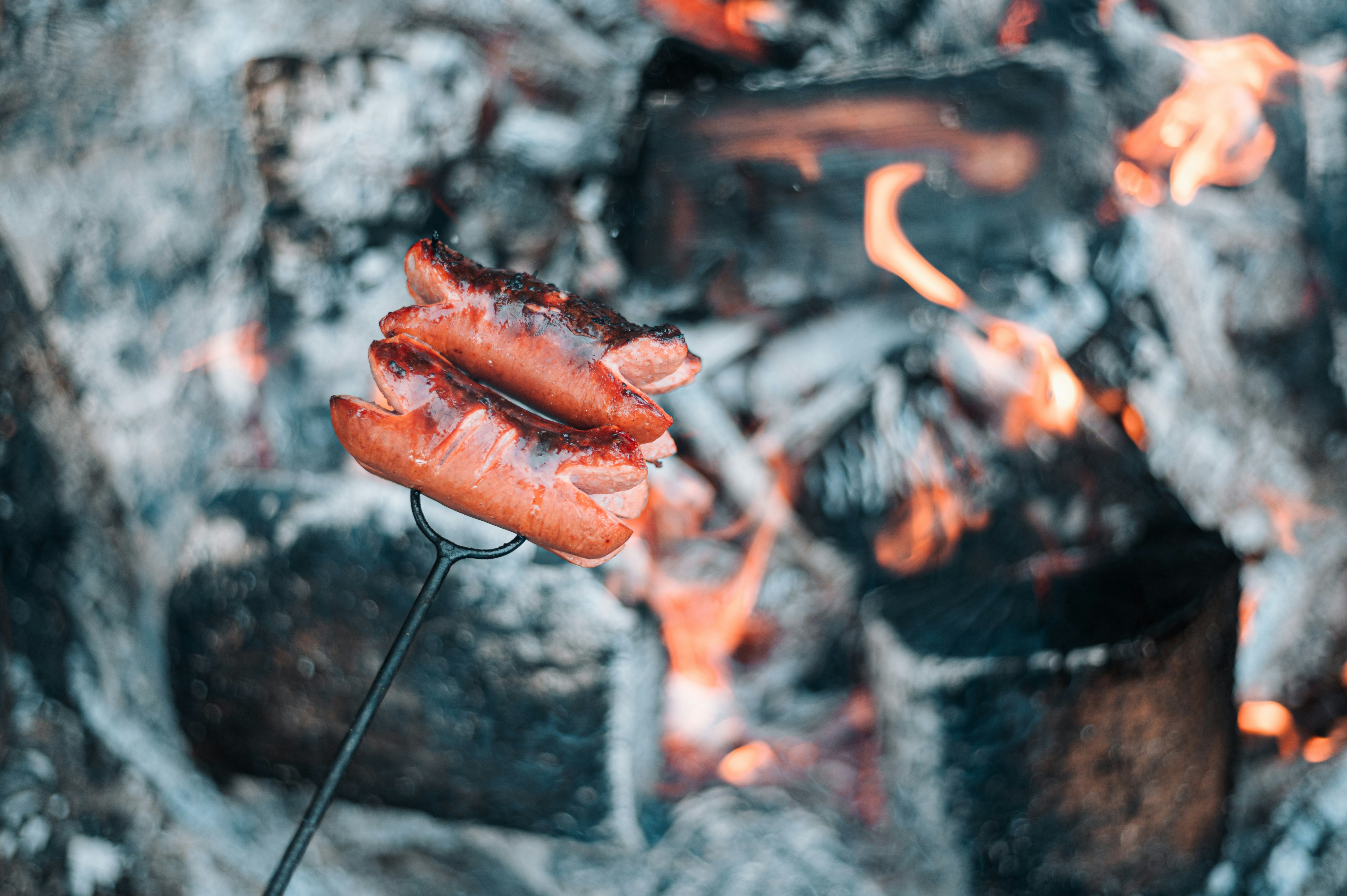 Grilled sausages on a skewer, held above a vibrant campfire with glowing embers in the background.