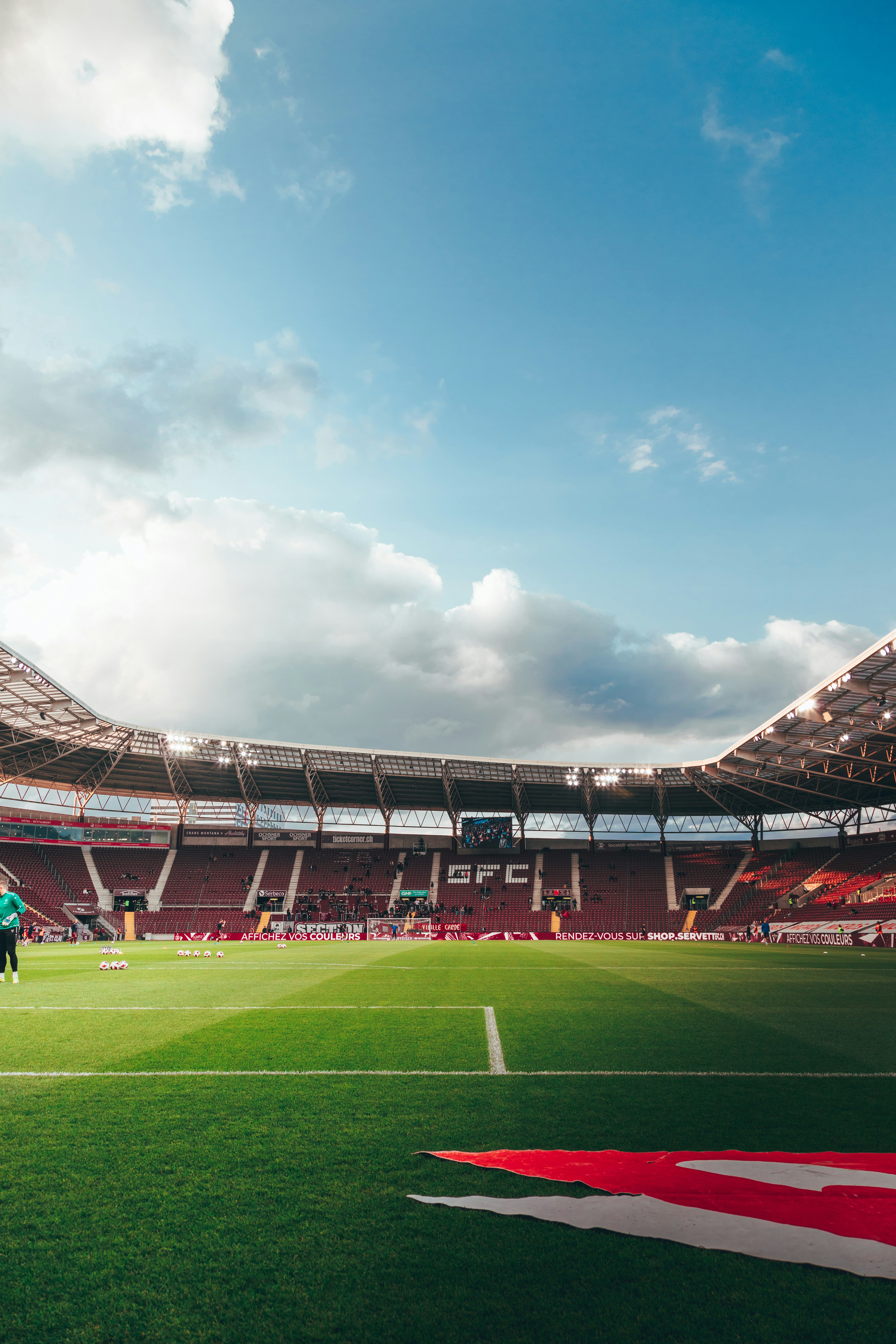 Vibrant stadium scene with lush green grass and a clear sky, showcasing preparations for an upcoming match. The stands are filled with anticipation.