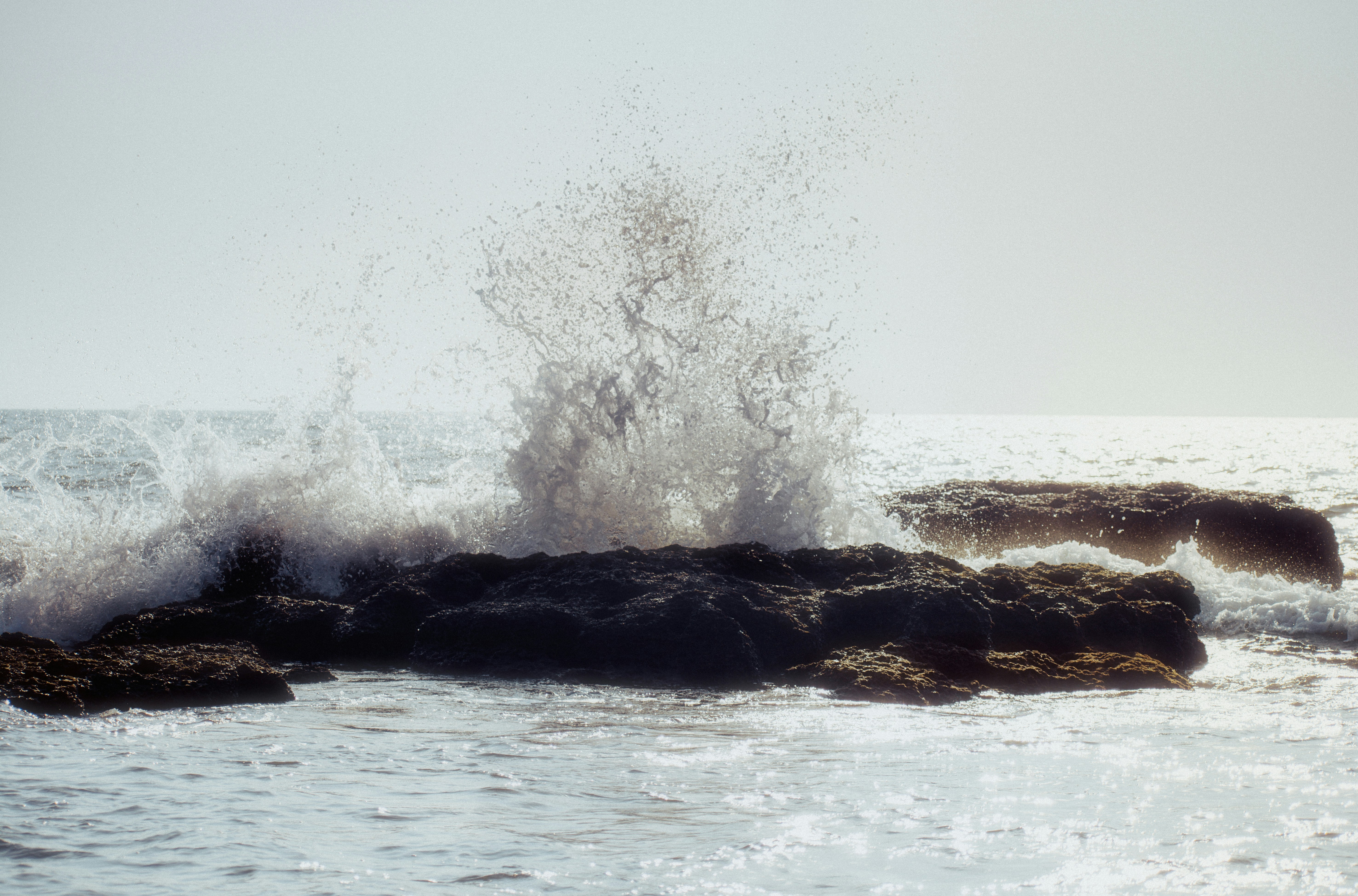 Ocean waves crashing against rocky outcrops, sending sprays of water into the air under soft sunlight.