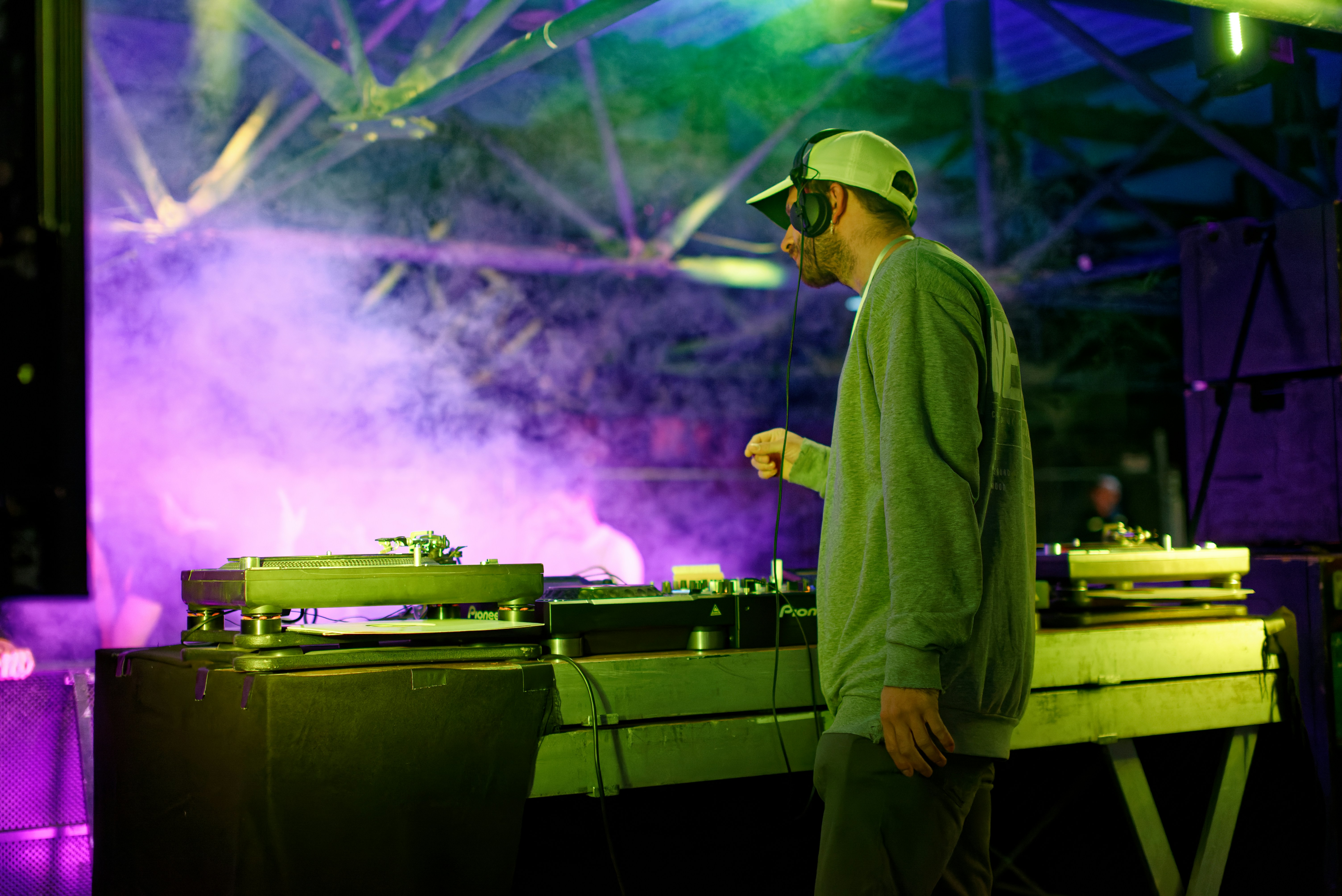 DJ performing at an outdoor event, surrounded by colorful lights and smoke. The atmosphere is energetic and immersive.