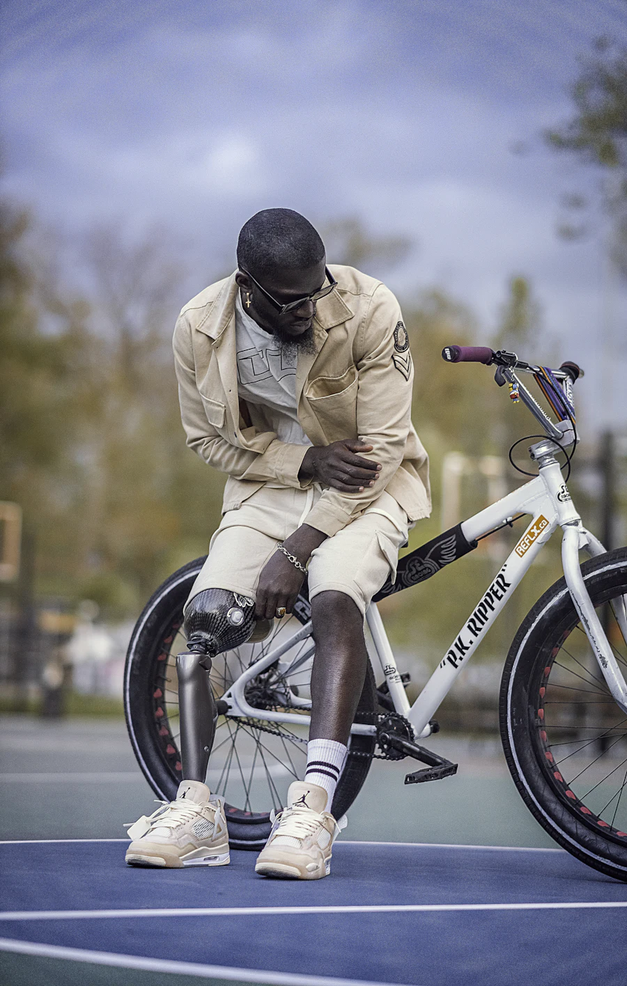 A man in beige clothing sits on a BMX bike, leaning forward and looking down. The bike’s white frame and colorful handlebars stand out against a soft blue sky and blurred trees.