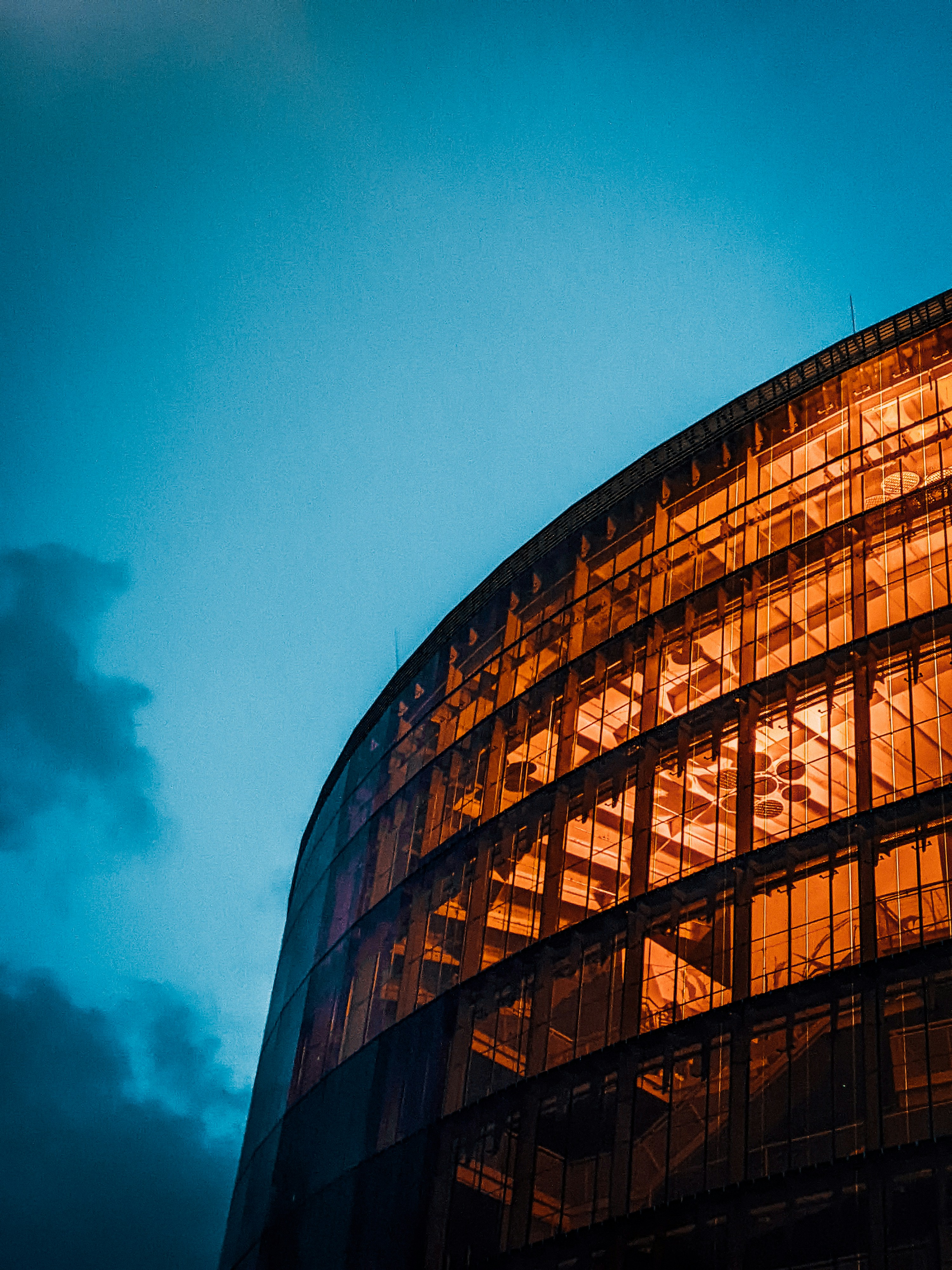 Curved glass office tower glows orange from interior lighting against a deep blue evening sky. The architectural grid and reflections emphasize the building's curvature.