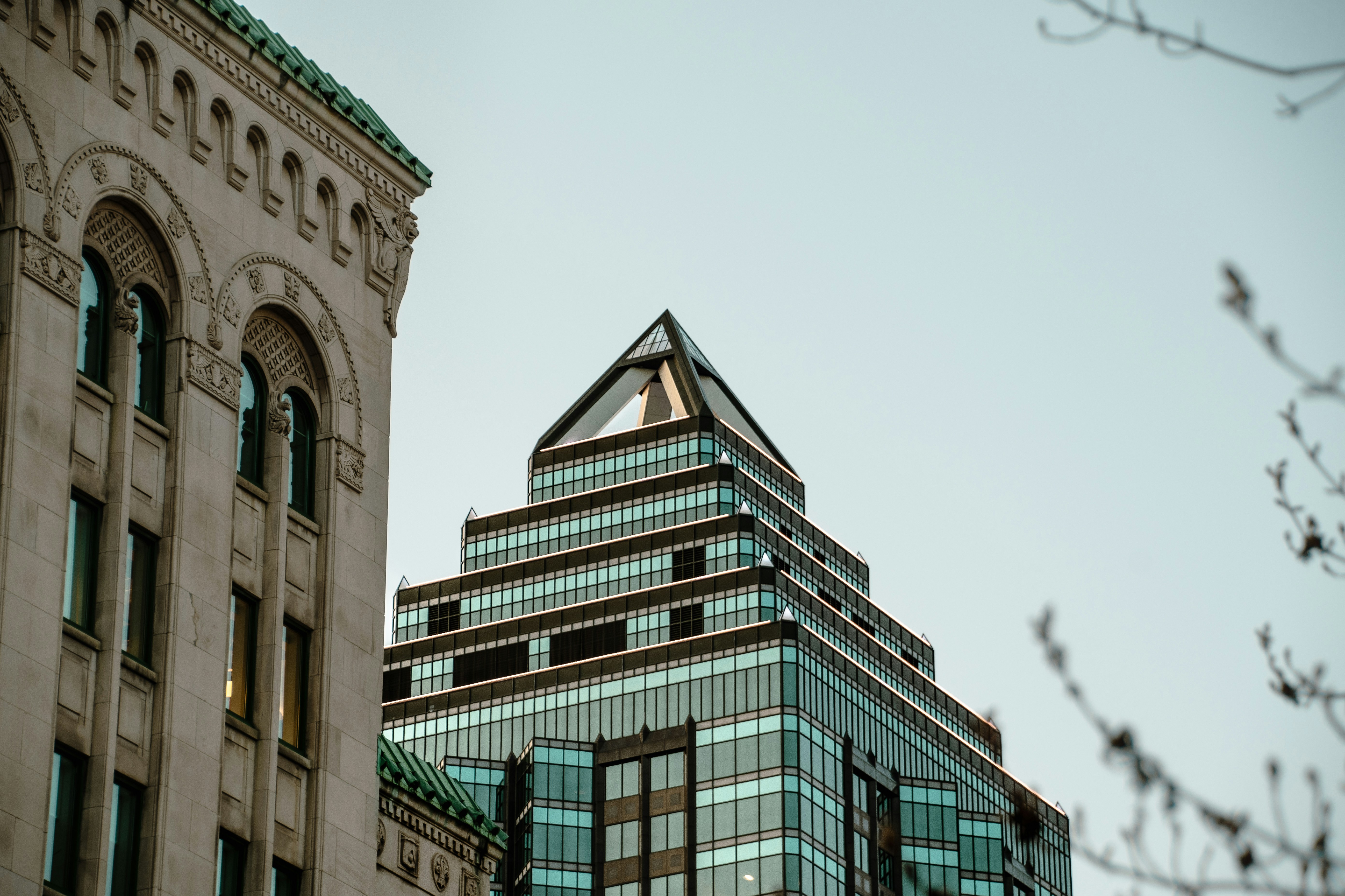 Modern glass skyscraper with distinct triangular top framed by older stone building and bare branches against the sky.