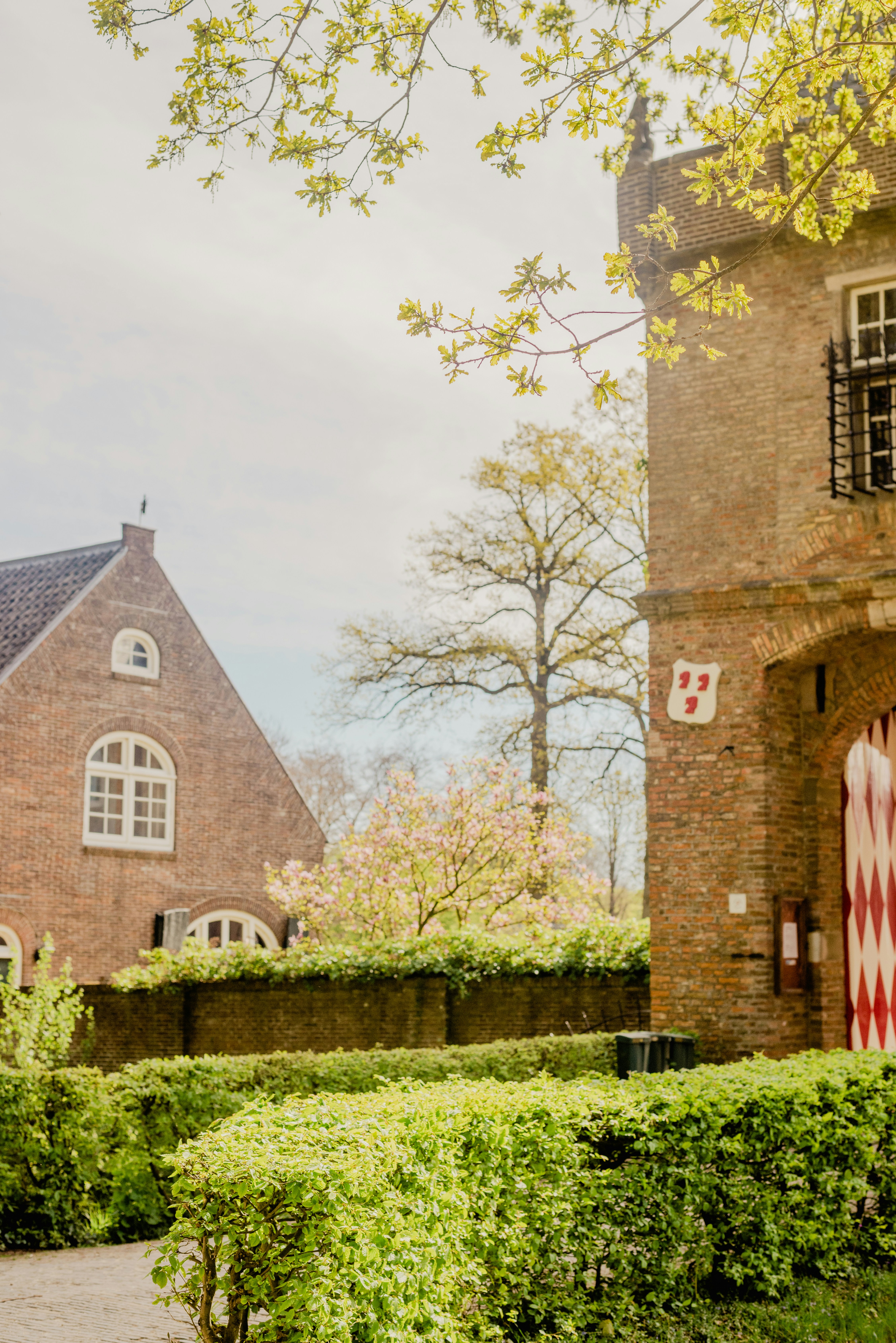 Historic brick buildings surrounded by lush green hedges and blossoming trees under a clear sky.