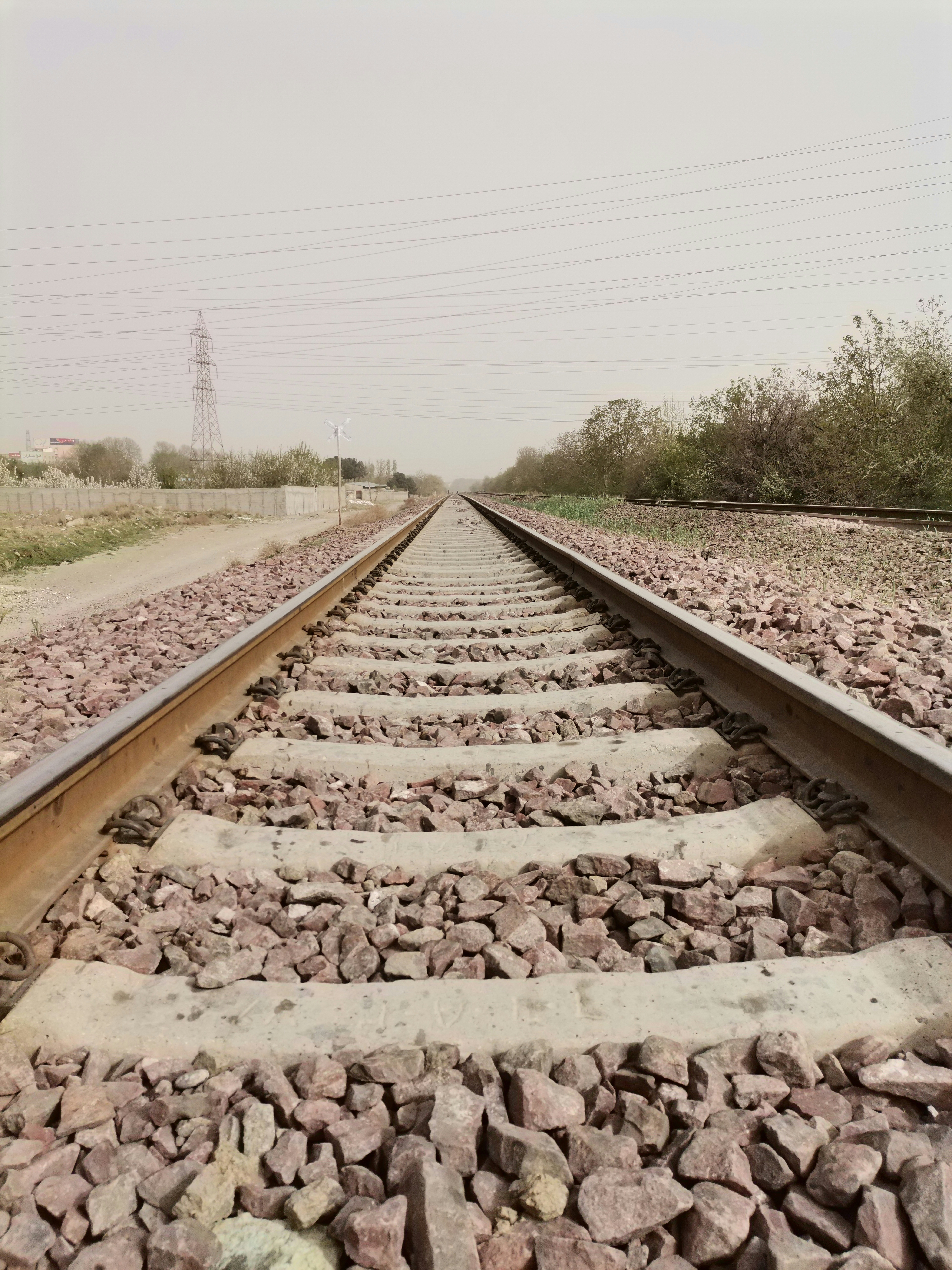 Low-angle view along parallel railroad tracks receding into a hazy horizon, with loose ballast and distant utility lines creating a vanishing point.
