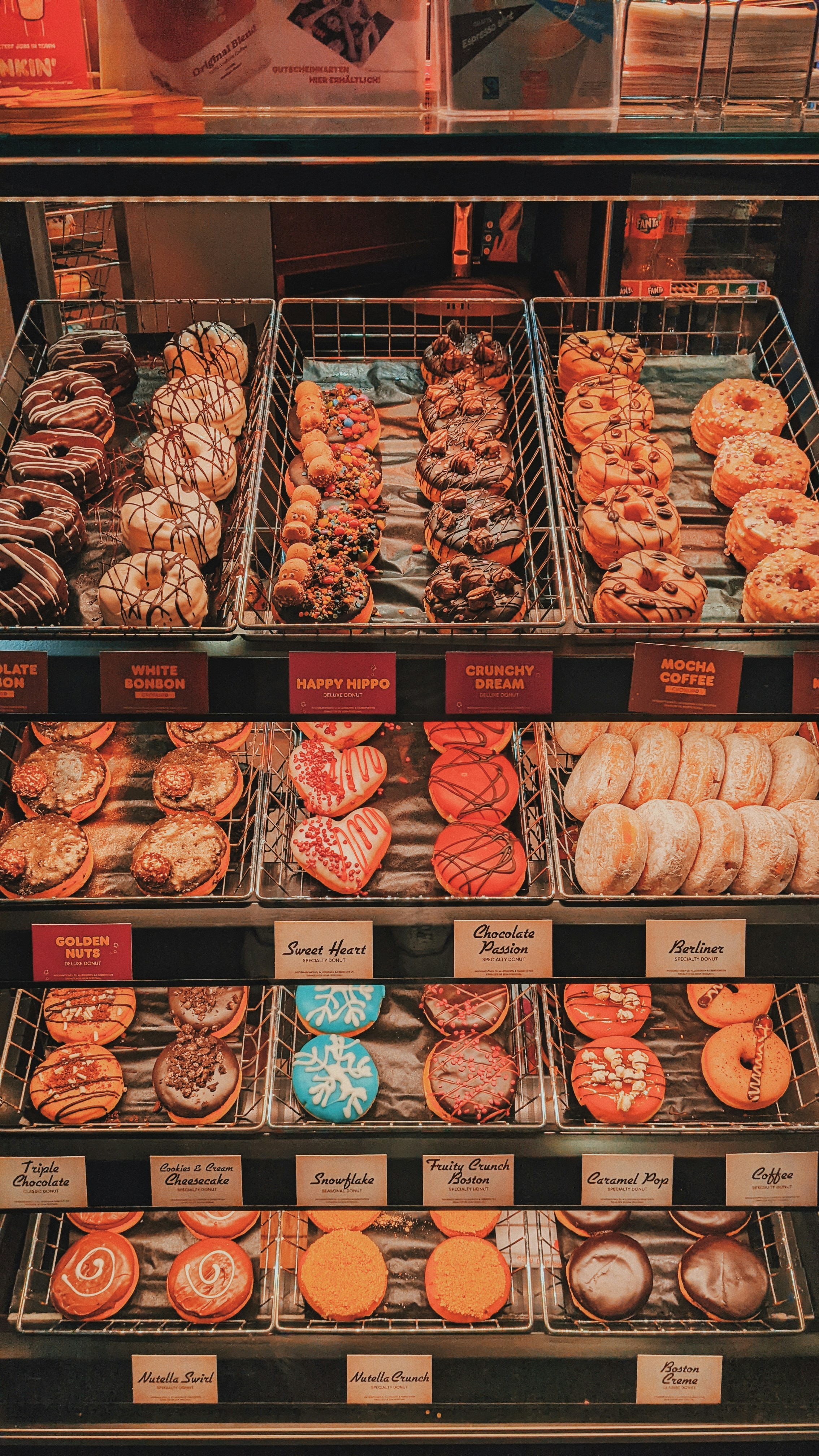 Close-up photograph of a bakery display case filled with assorted donuts arranged in metal trays under warm lighting.