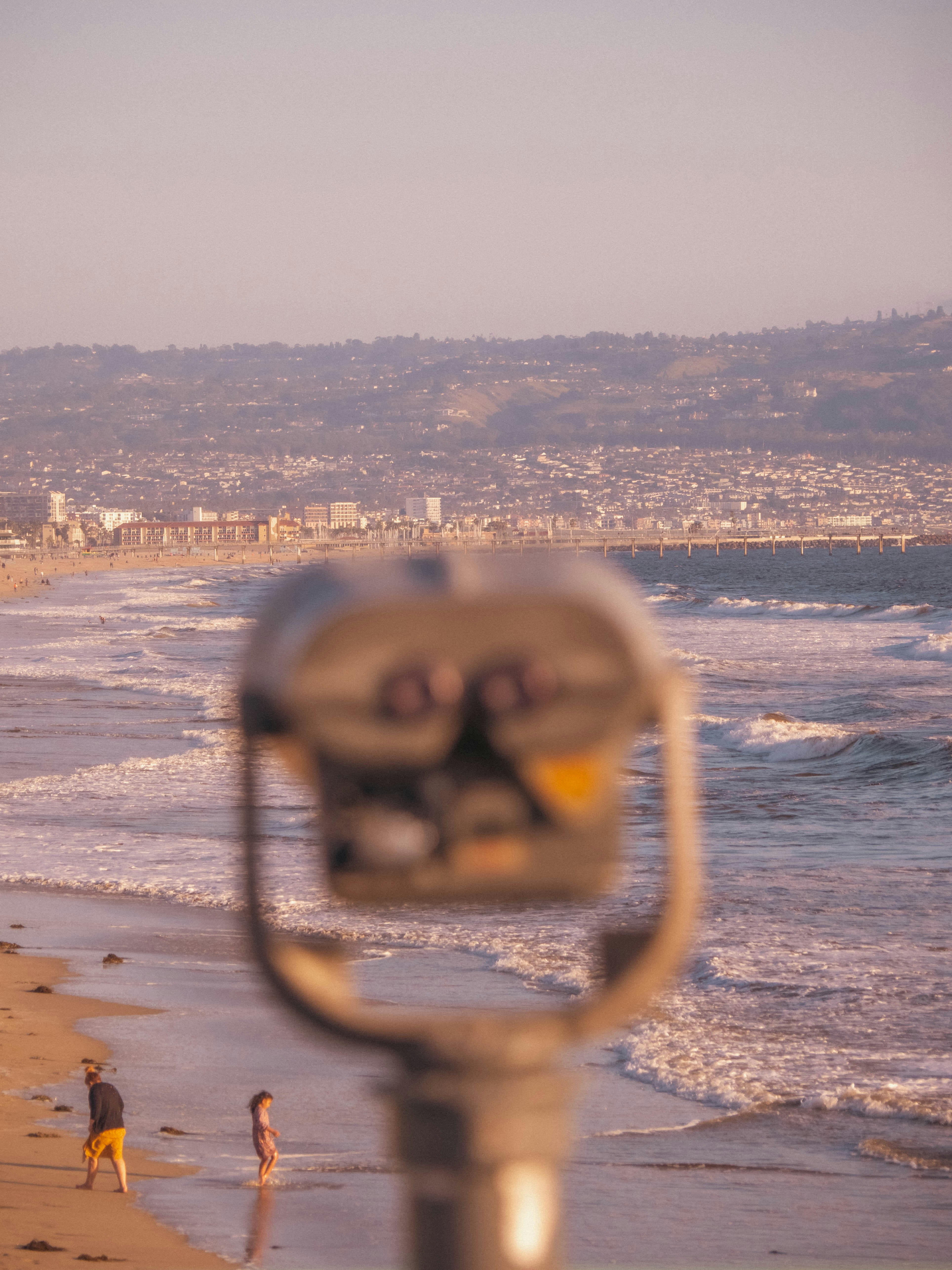 A beachside coin-operated viewer stands in focus, framing a blurred view of the sandy shore and distant cityscape. Waves gently lap at the beach, creating a serene atmosphere.