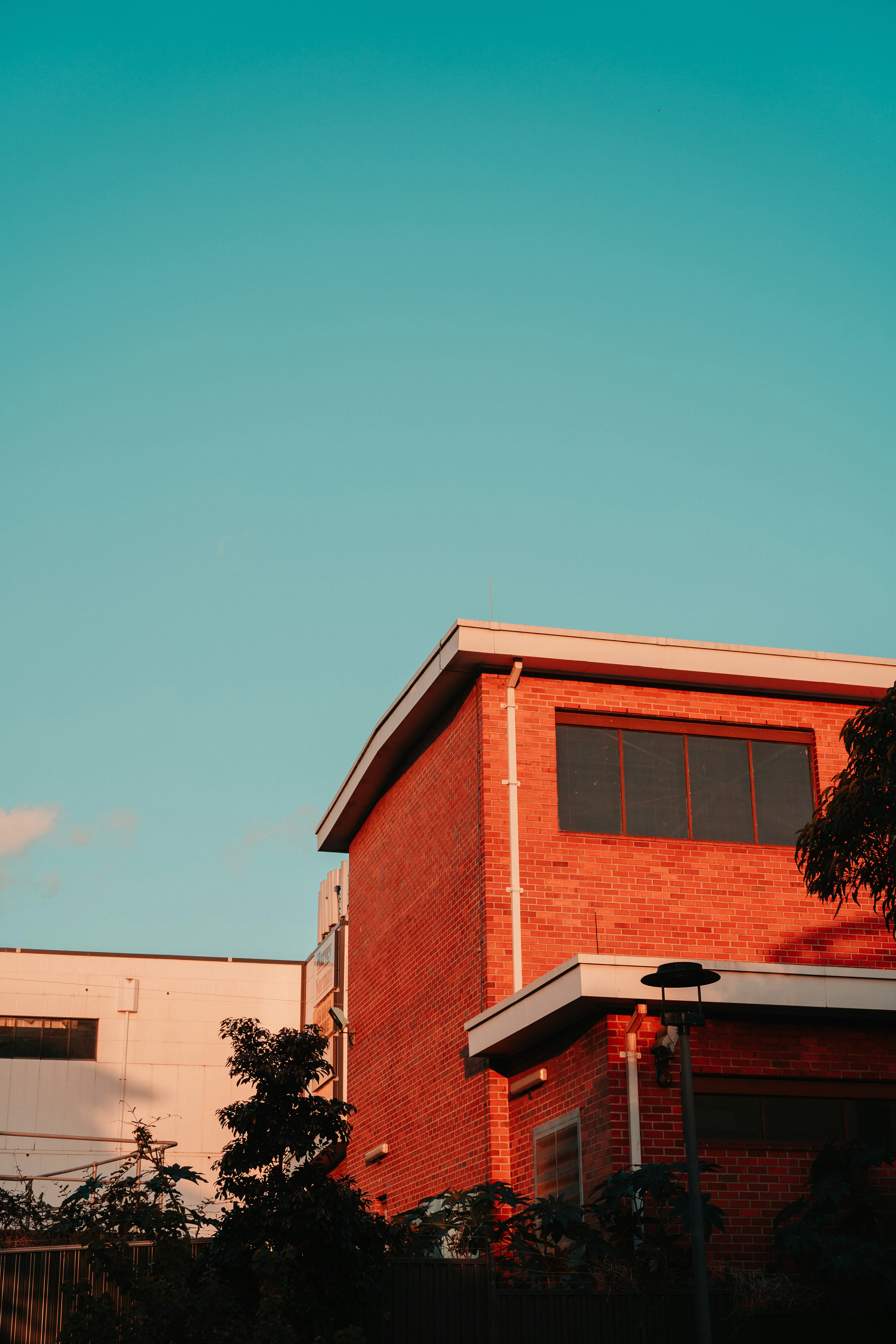 Brick building with large windows and a sloped roof, framed by greenery against a clear blue sky.