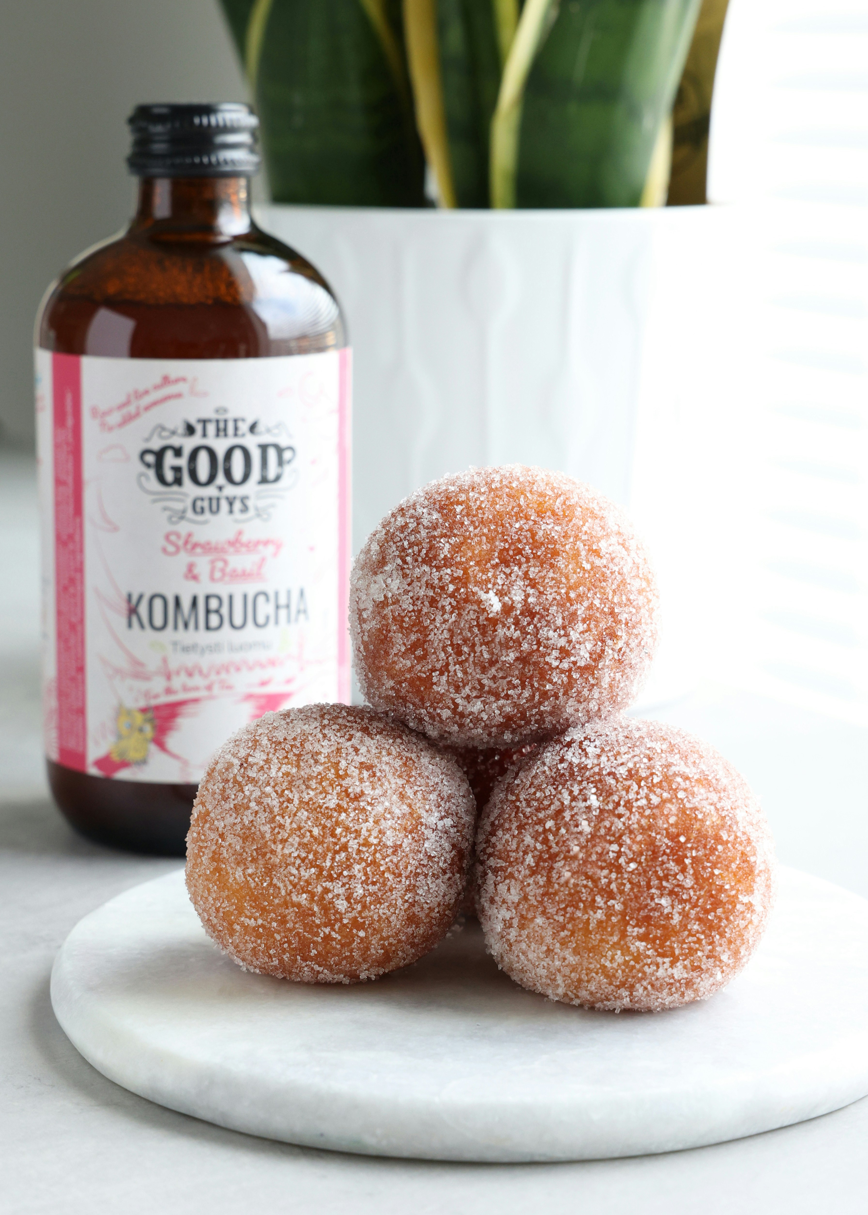 Three sugar-coated doughnuts stacked on a marble surface, accompanied by a bottle of strawberry and basil kombucha. A vibrant green plant adds a touch of freshness.