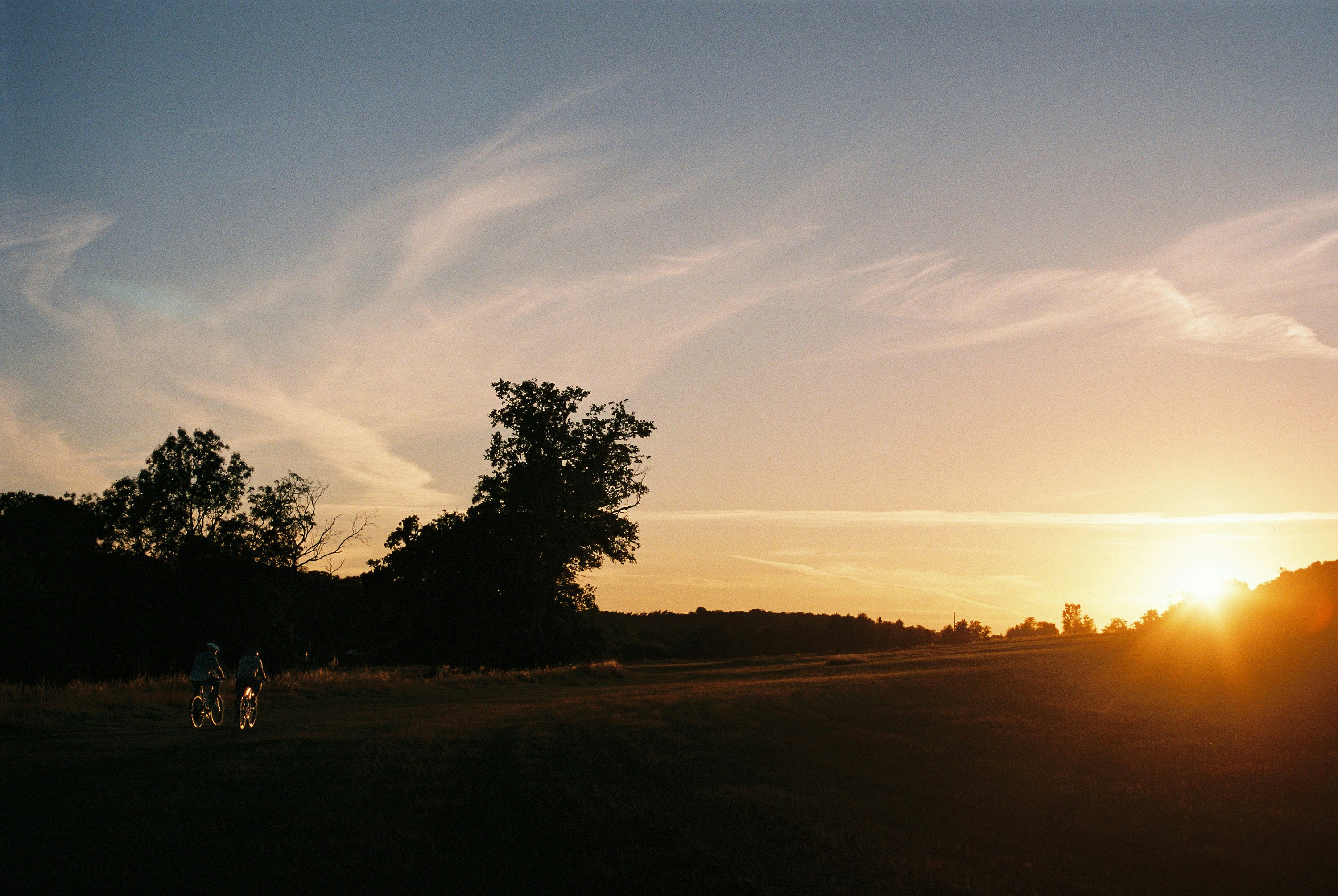Two cyclists navigate a tranquil landscape as the sun sets behind distant trees, casting a warm glow across the field.