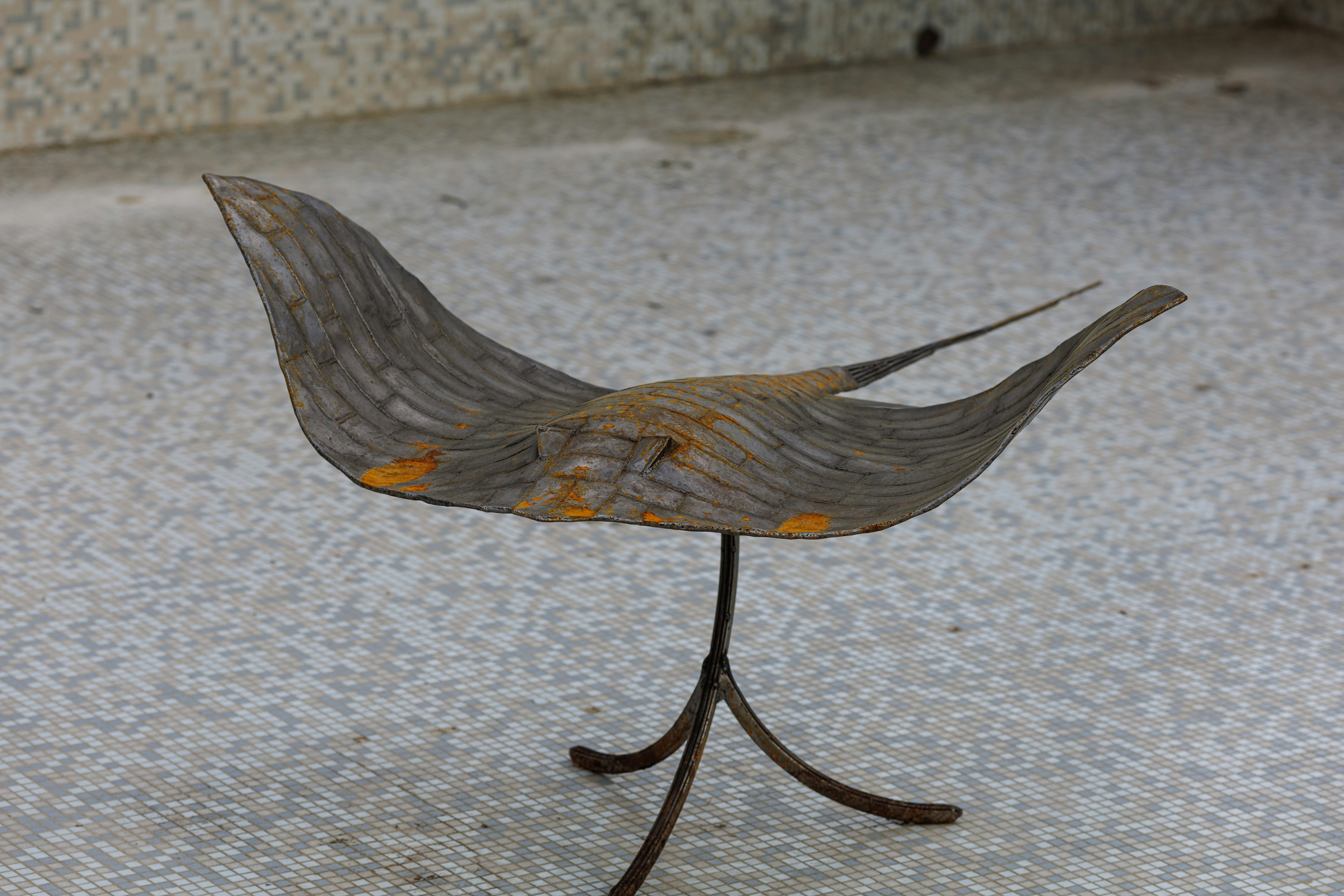 Abstract metal sculpture resembling a bird in flight, resting on a delicate stand. The piece features intricate textures and a weathered finish.
