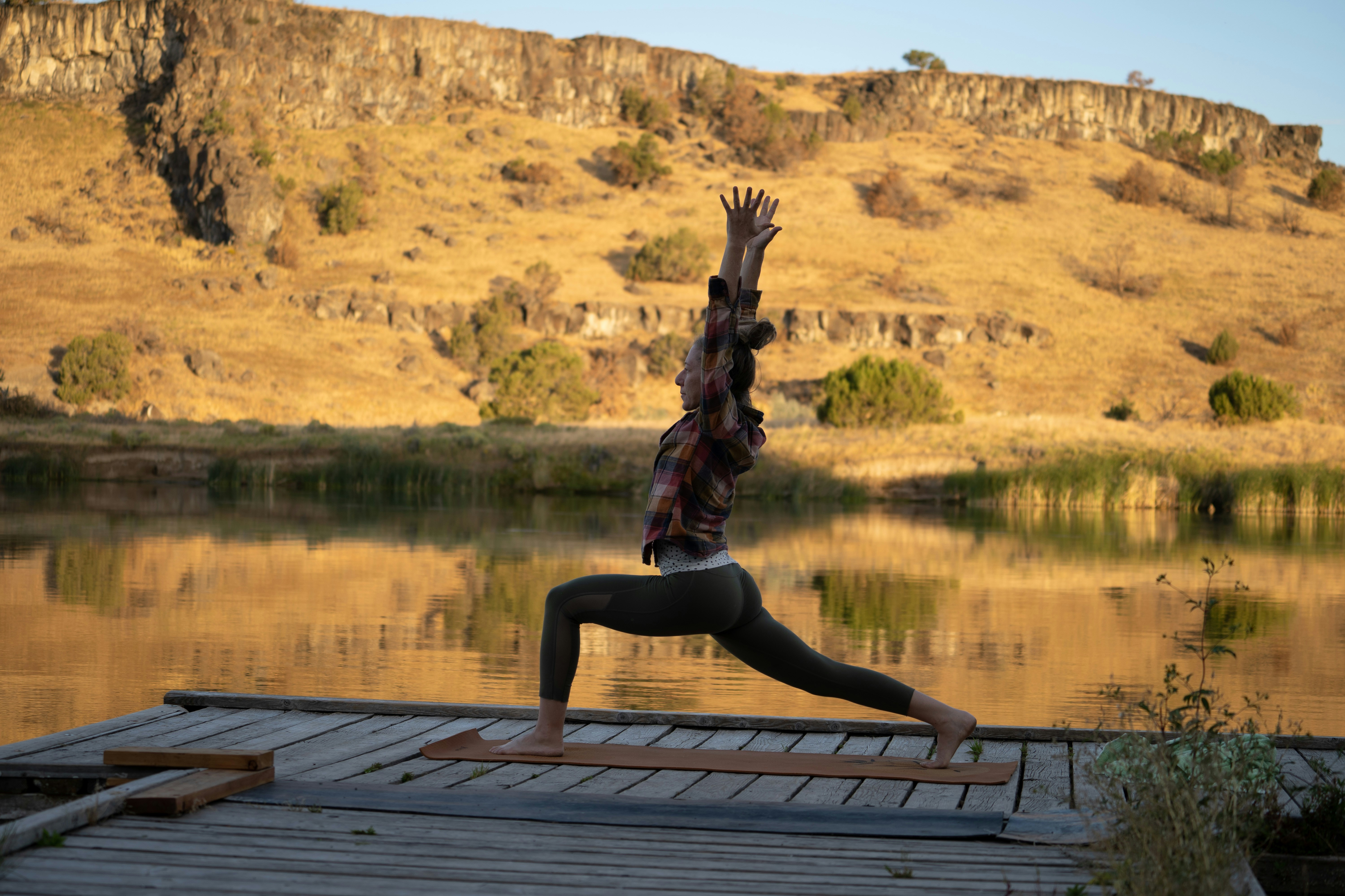 A woman practicing yoga in a warrior pose on a wooden dock, with tranquil waters and golden hills reflecting the serene atmosphere.