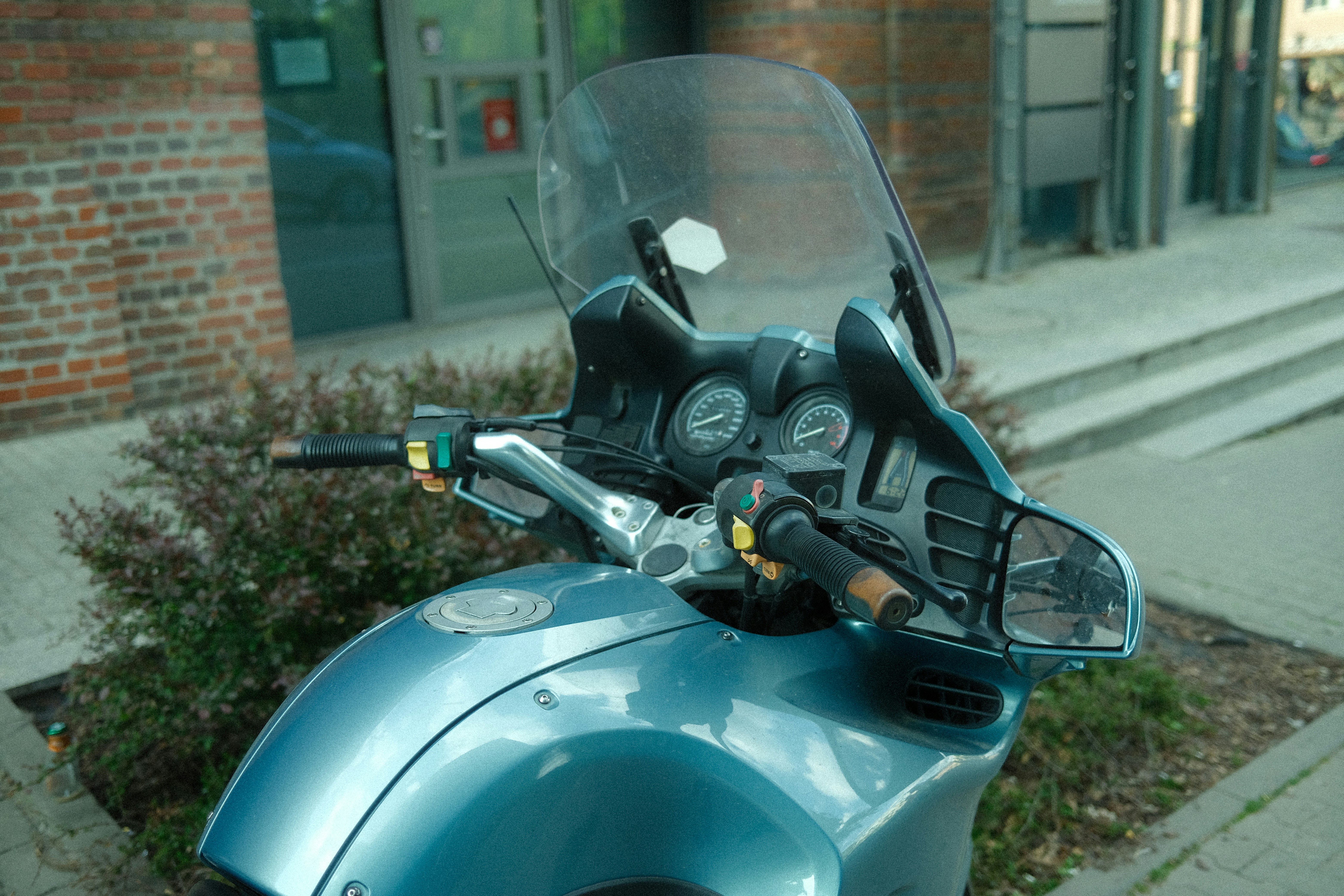 Close-up view of a motorcycle's dashboard and handlebars, highlighting the controls and gauges. The setting features a brick wall backdrop and greenery.