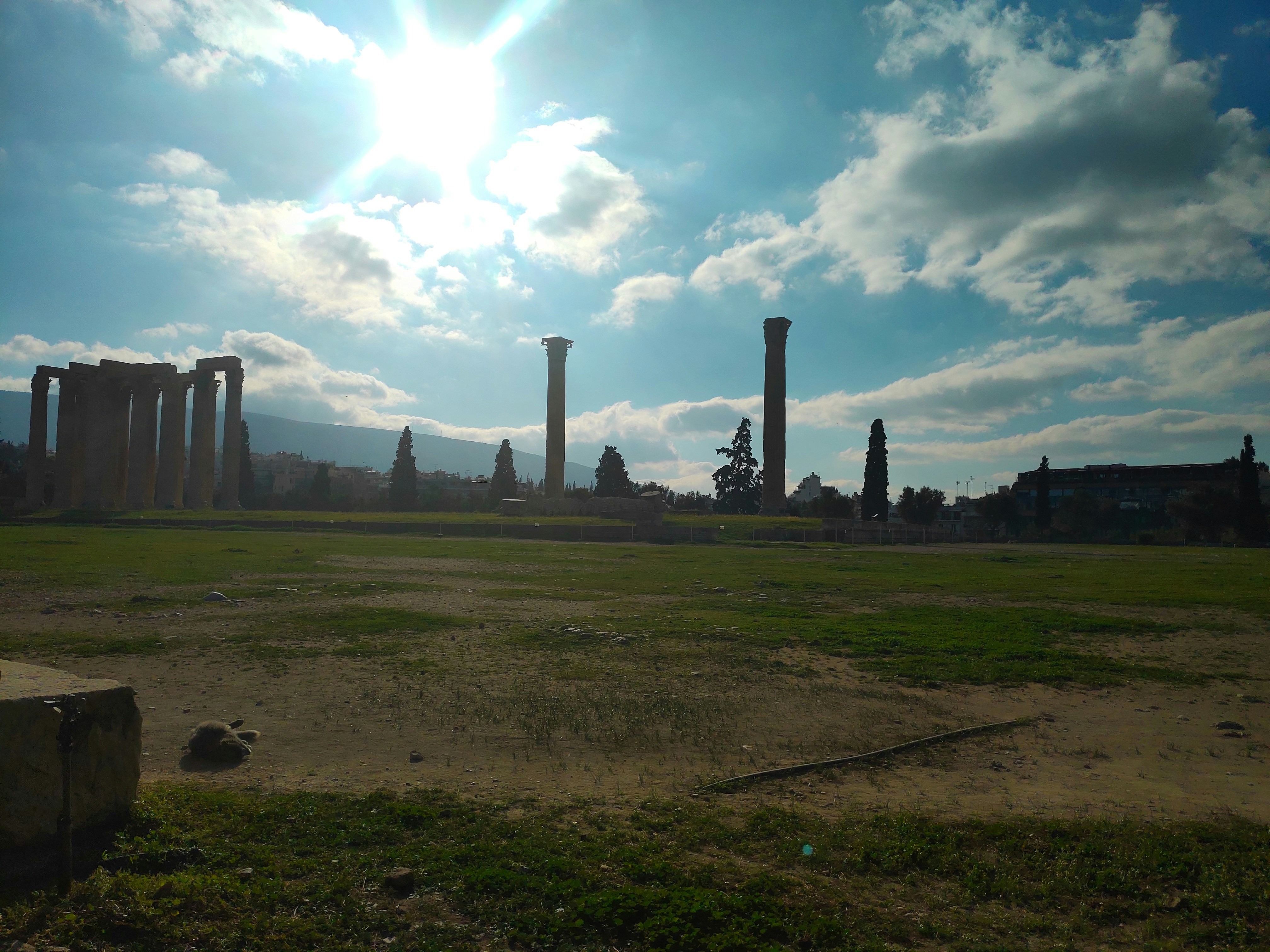 Ancient columns and ruins rise across a grassy field beneath a bright, sunlit sky. A few tall pillars remain as remnants of a historic site.