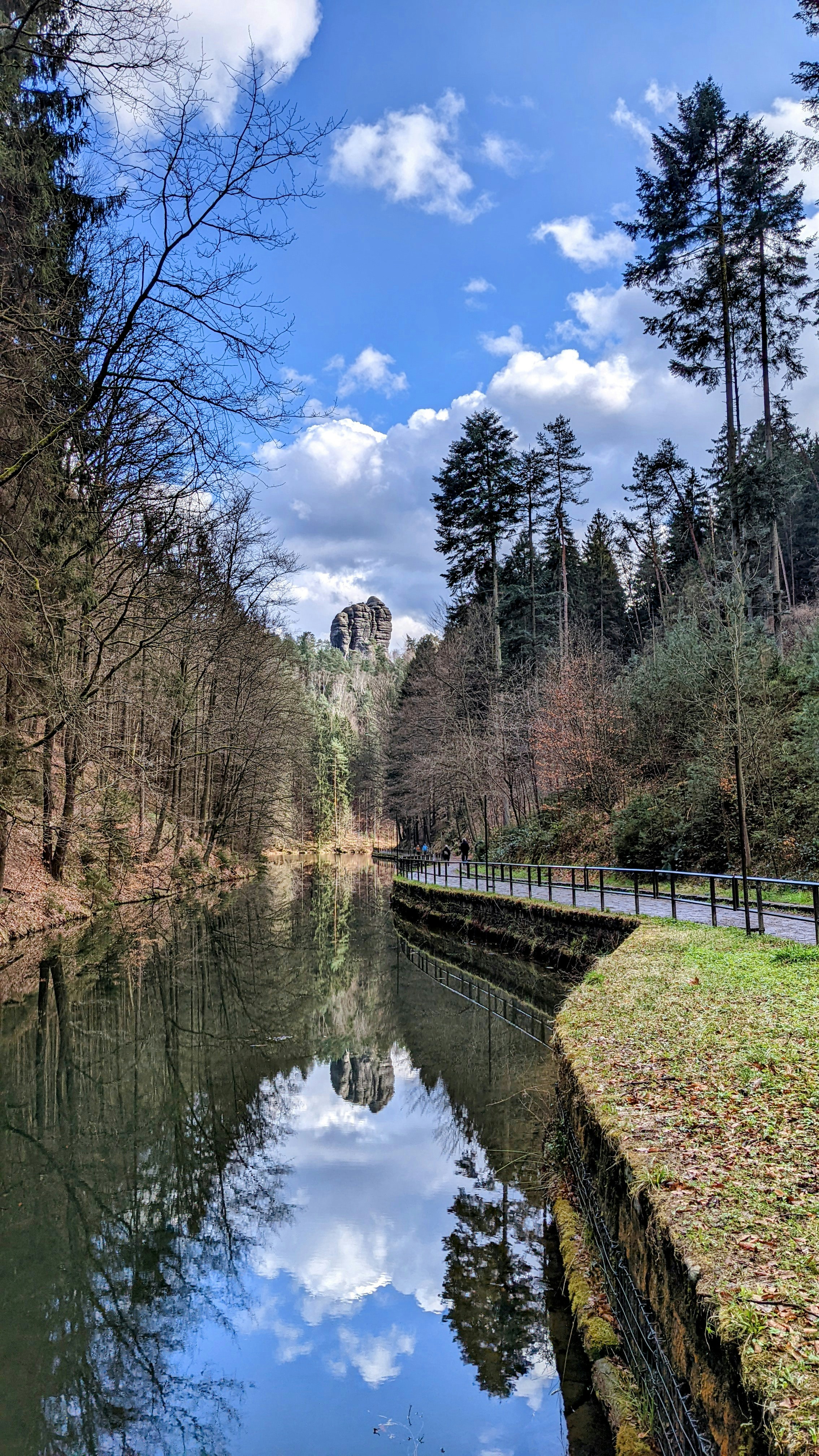 Serene river reflecting towering rock formations and a vibrant sky, surrounded by tall trees in early spring.