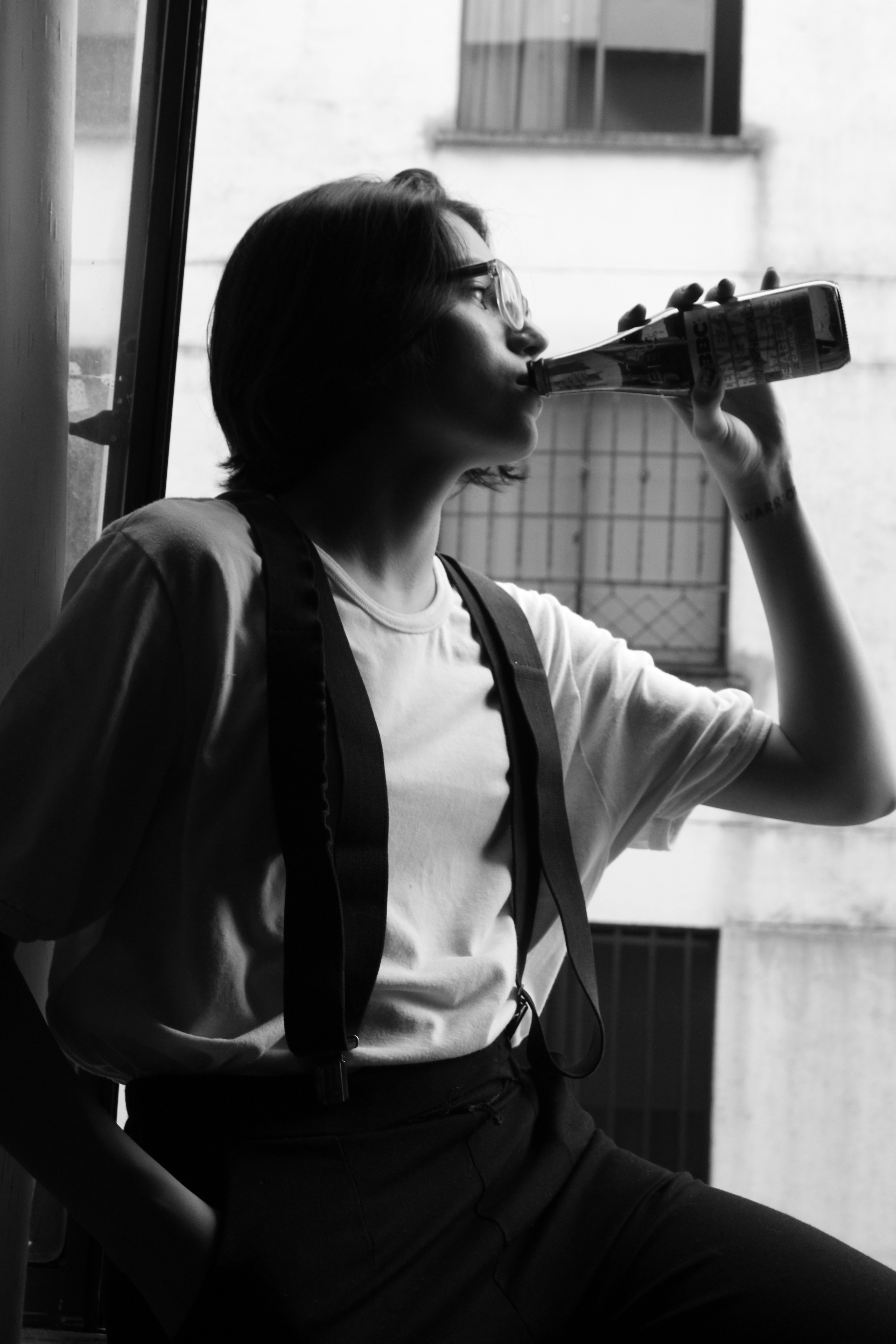 Individual sipping from a bottle while seated by a window, framed in monochrome tones. The scene captures an intimate moment of pause amid an urban backdrop.