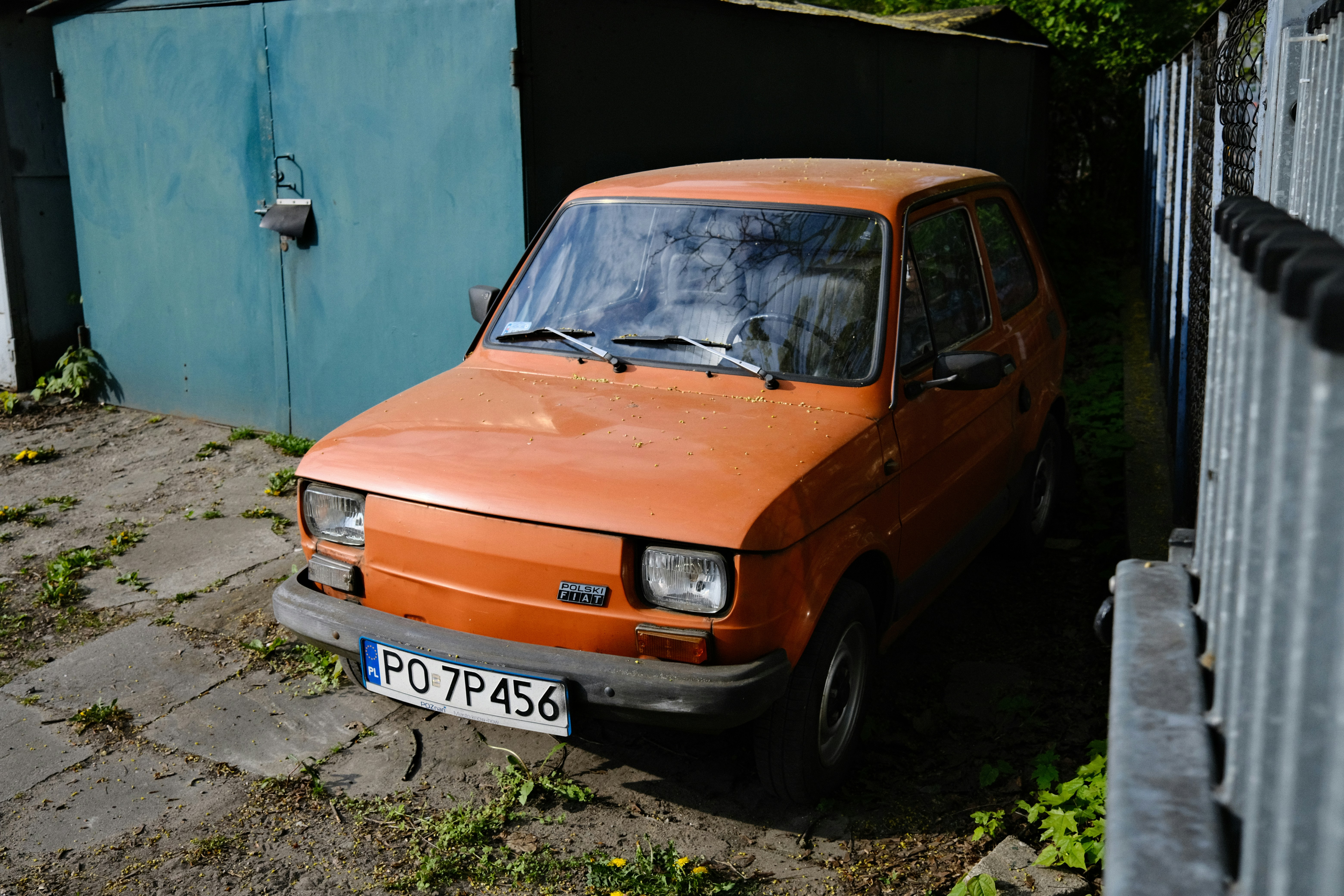 An orange vintage car parked beside a green shed, partially obscured by foliage and a fence. The scene evokes a sense of nostalgia and abandonment.