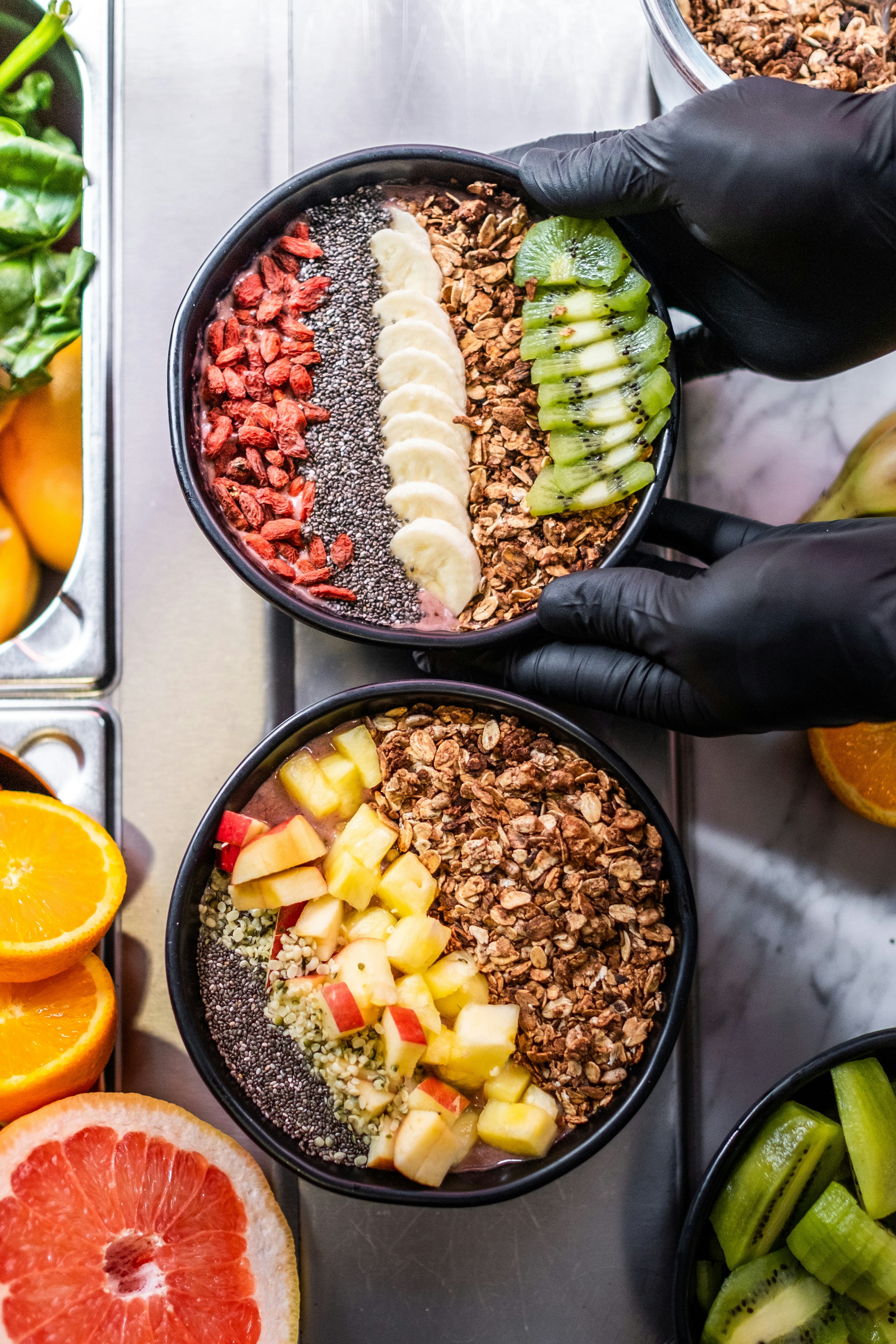 Gloved hands curate kiwi-banana granola bowls on a chrome prep line