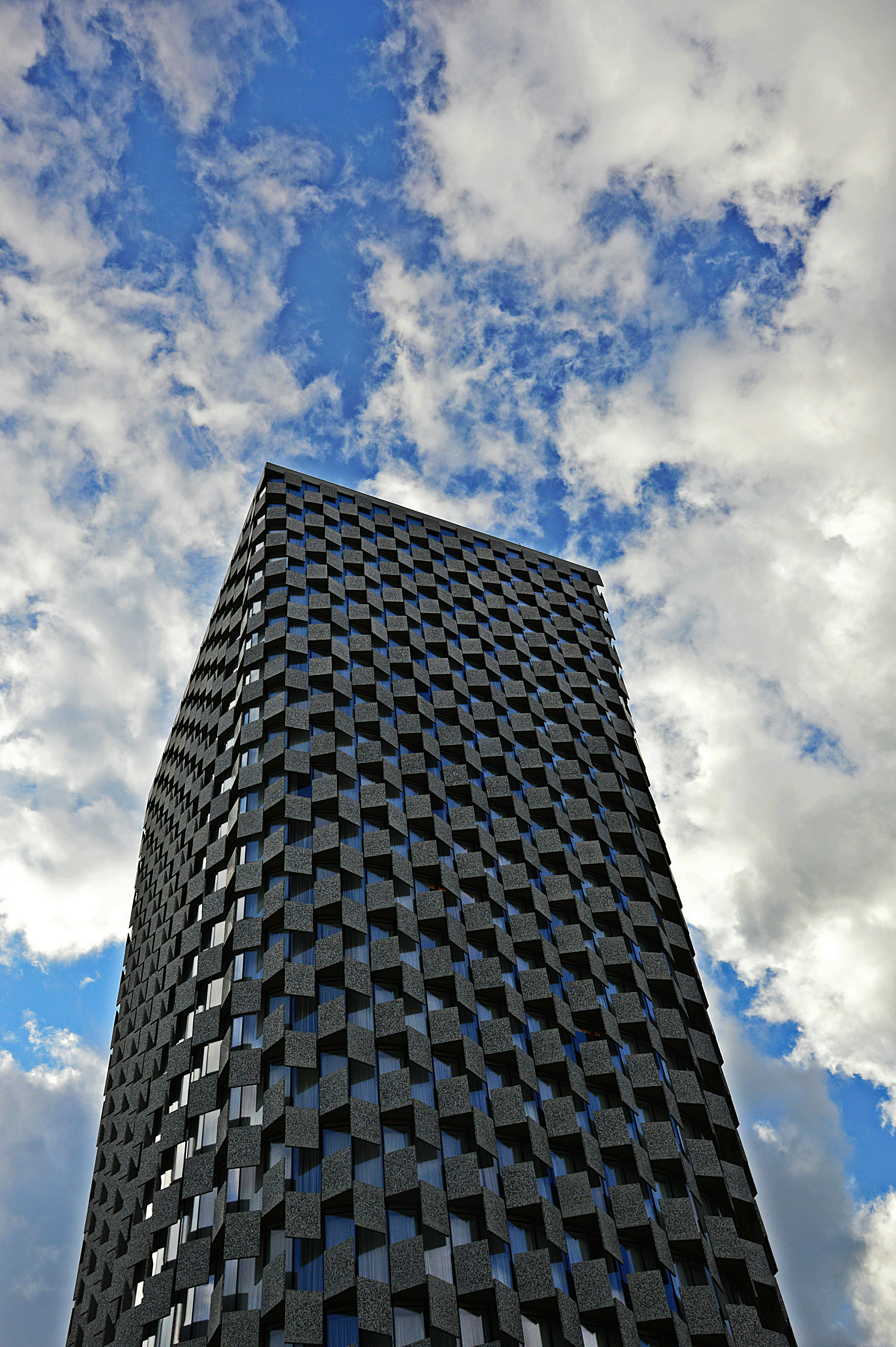 A striking modern building with a textured facade reaching towards a cloudy sky. The geometric patterns create a dynamic visual contrast.