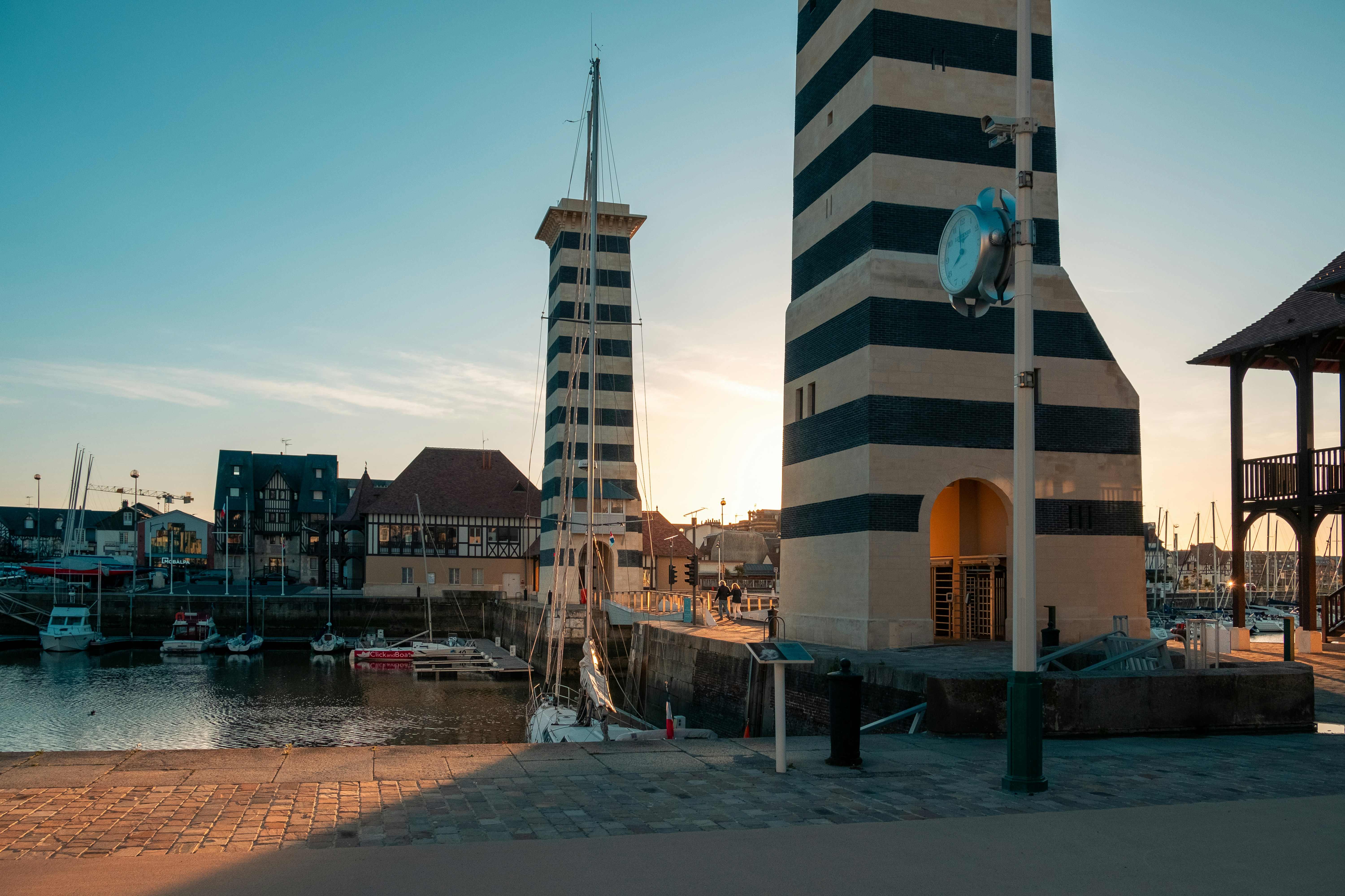 Lighthouses stand tall beside a tranquil harbor at sunset, with boats gently bobbing in the water. The scene evokes a sense of calm and maritime charm.