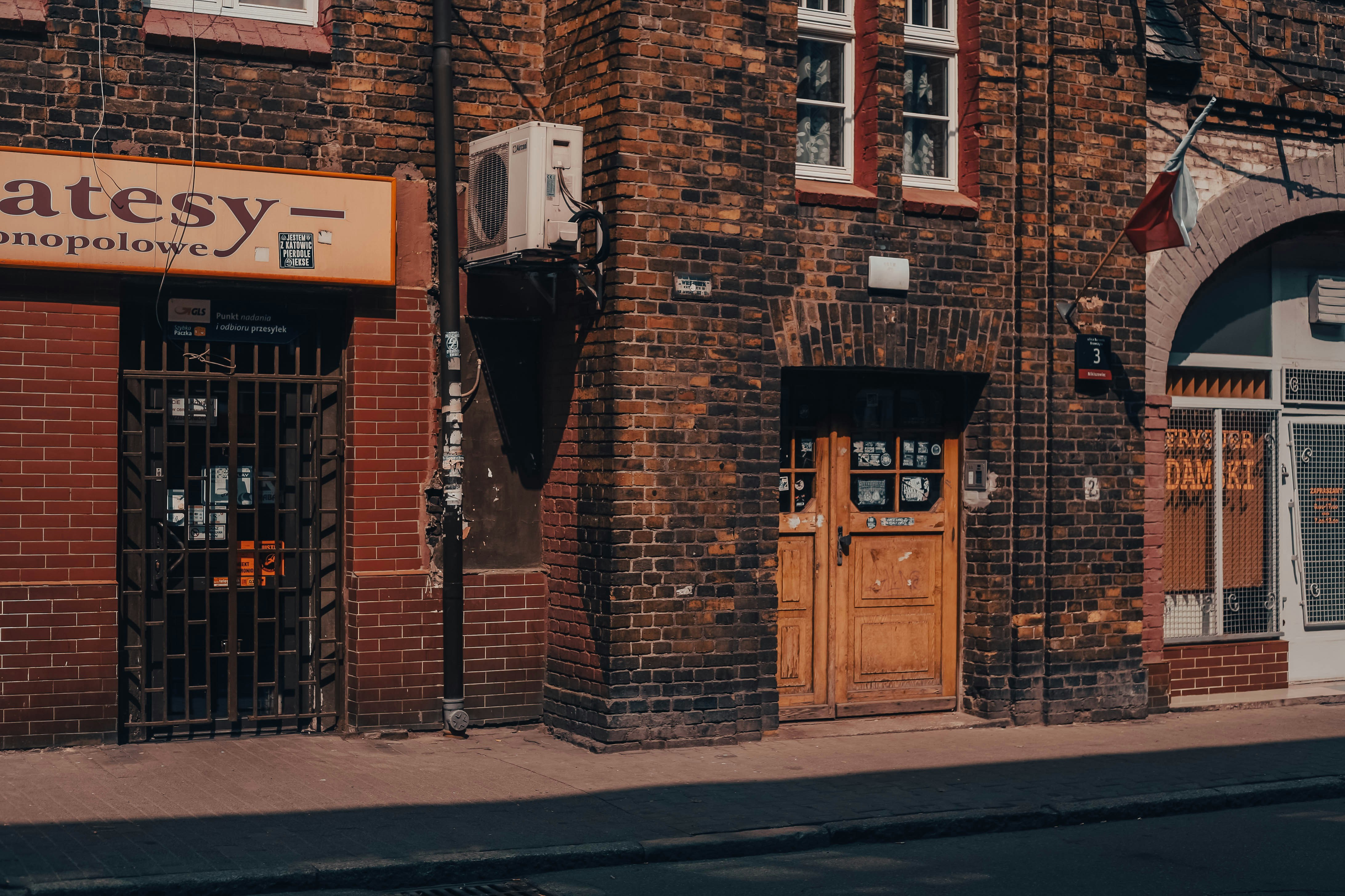Brick building with a wooden door and window, flanked by a shop and an archway, under warm sunlight.