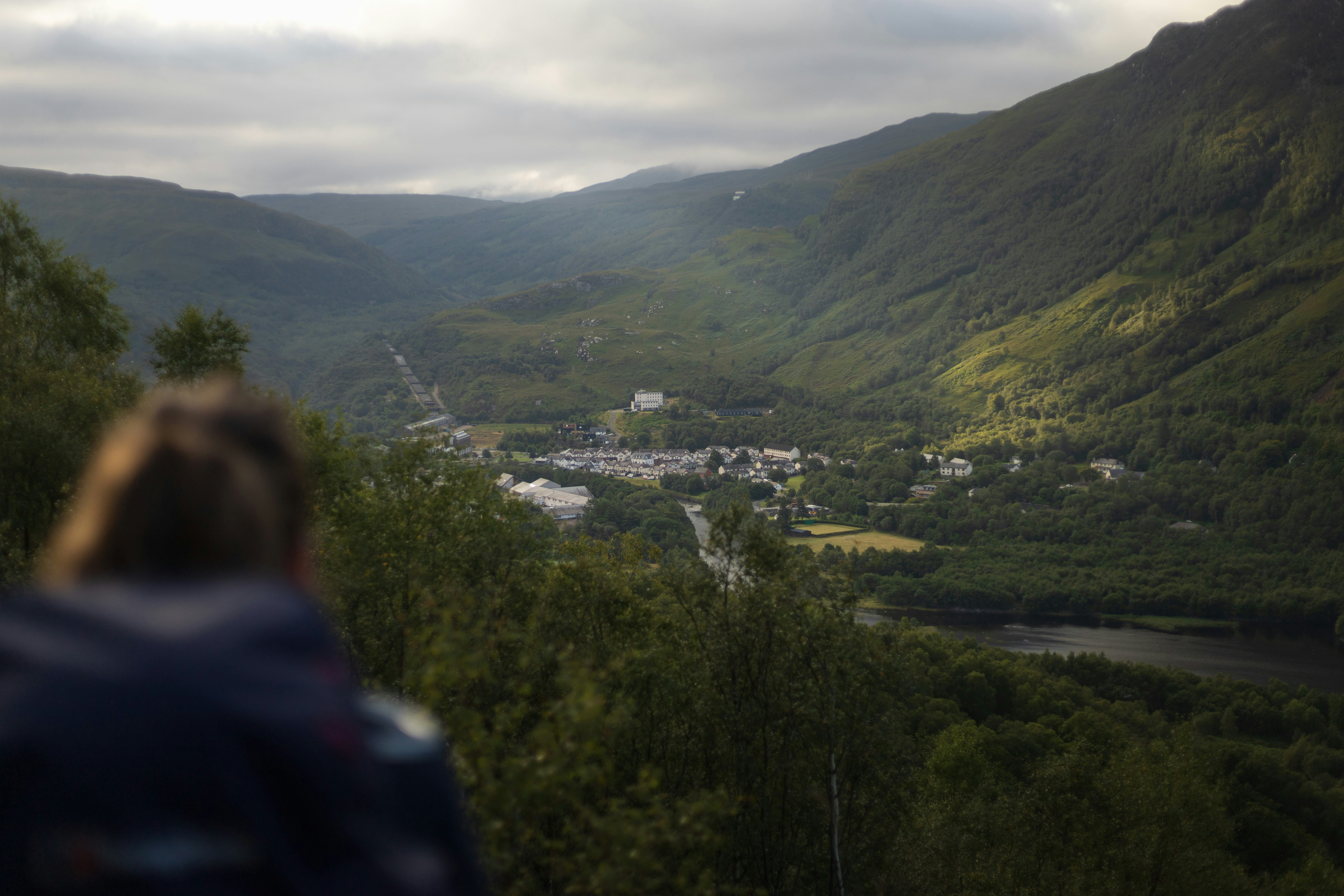 A hiker gazes over a lush valley, with a small town nestled among the hills and a river winding through the landscape. The soft light highlights the contours of the mountains.