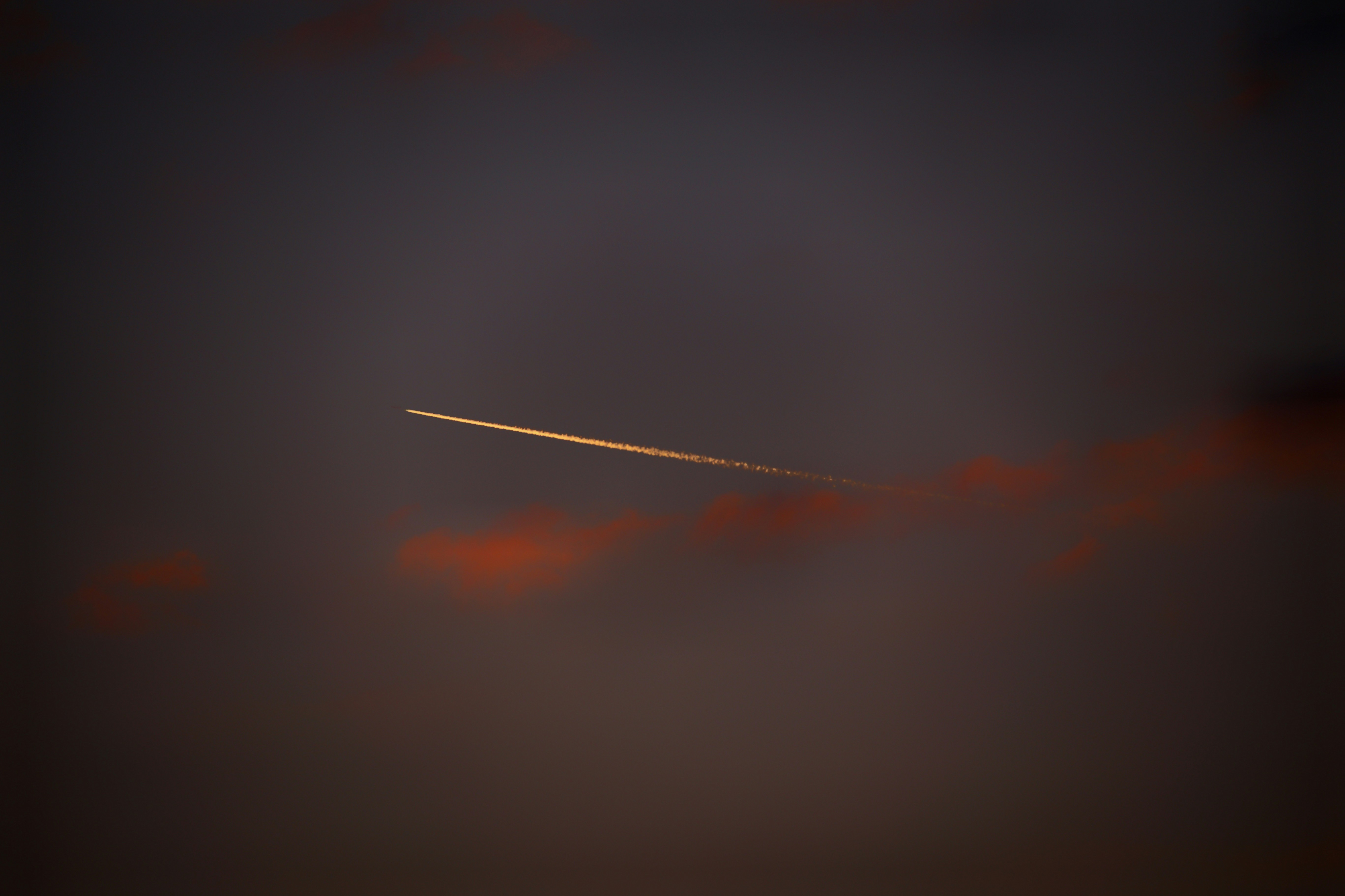 Contrail of an aircraft slicing through a twilight sky, illuminated by the fading light and surrounded by soft clouds.