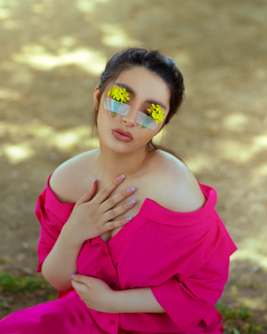 Woman in a bright pink off-shoulder outfit posing outdoors with yellow flowers placed over her eyes. Soft bokeh background and a hand resting on her chest create a stylized fashion portrait.
