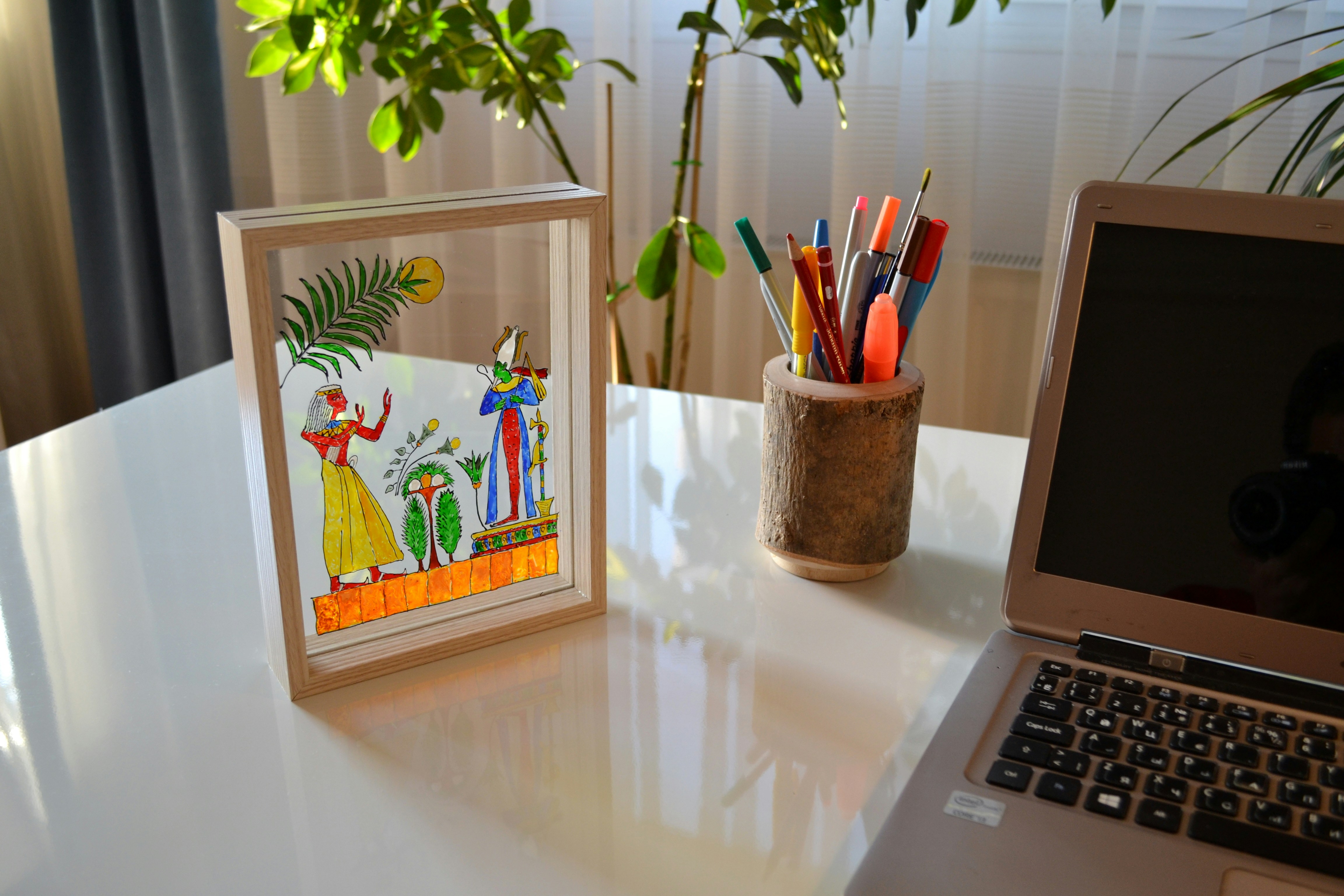Framed child's artwork on a bright desk beside a laptop and pencil jar