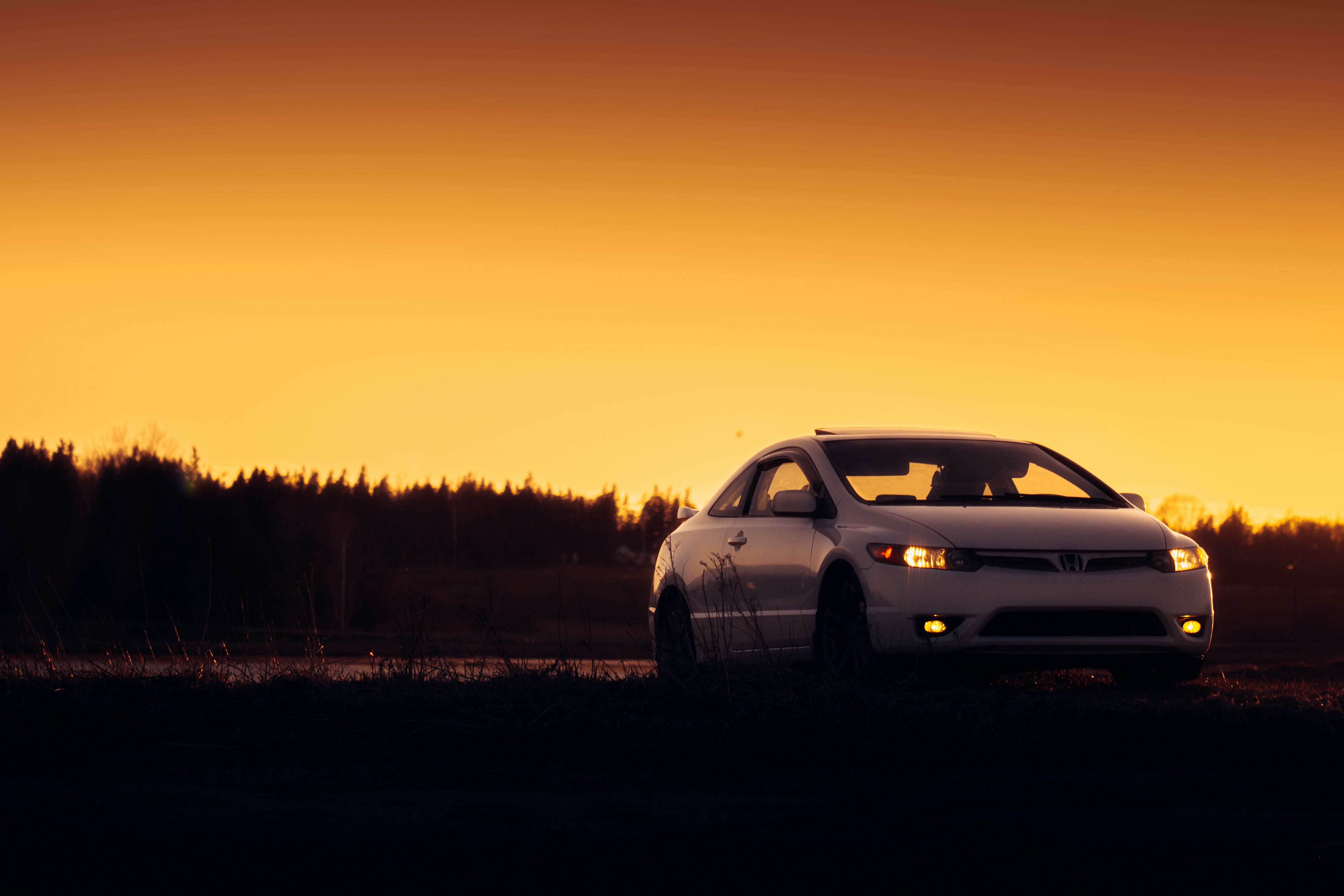 White car parked on a dark road, silhouetted against a vibrant sunset sky with gradient hues of orange and yellow.
