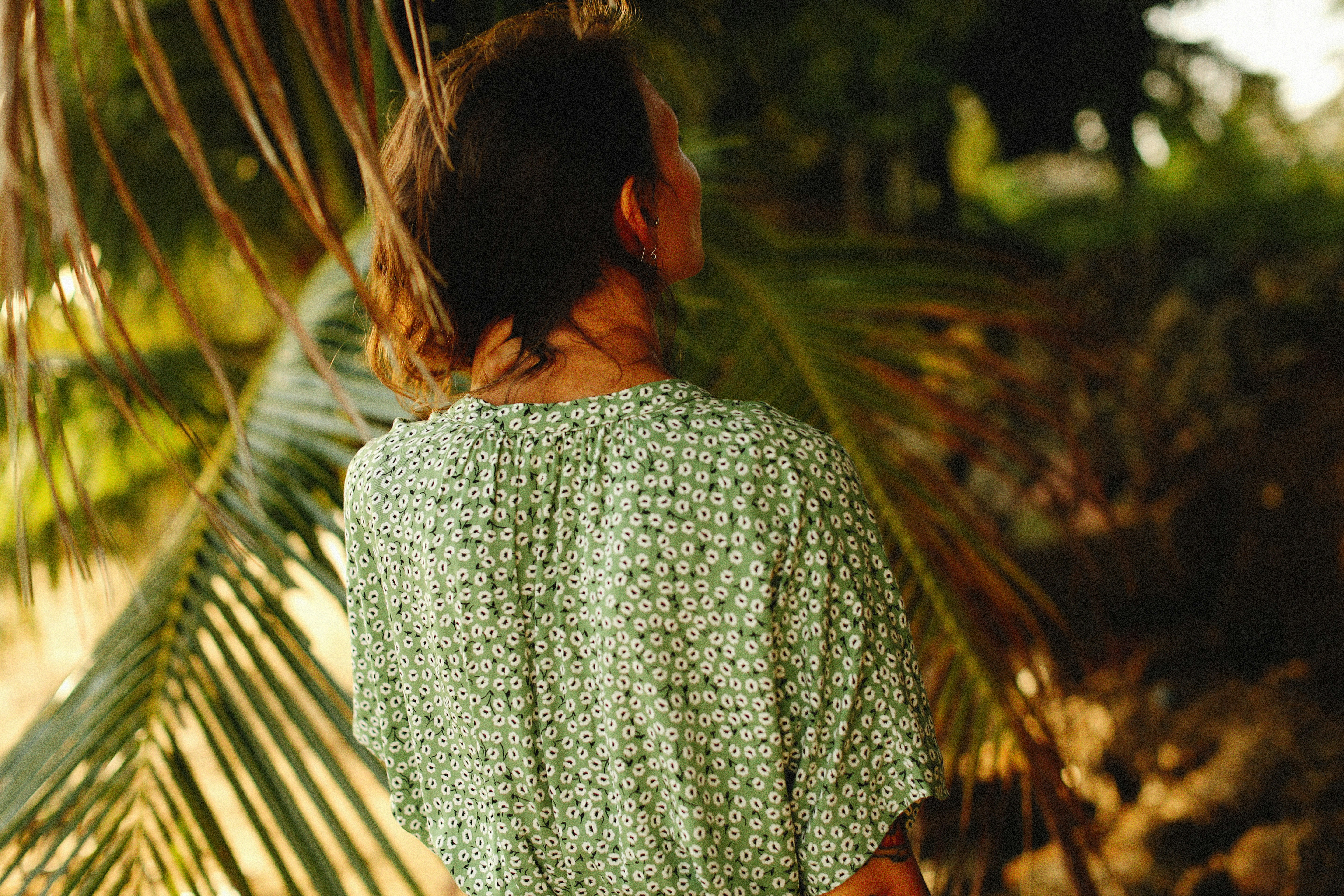 A woman in a green floral blouse stands with her back to the camera, framed by lush palm leaves, evoking a sense of tranquility in a natural setting.