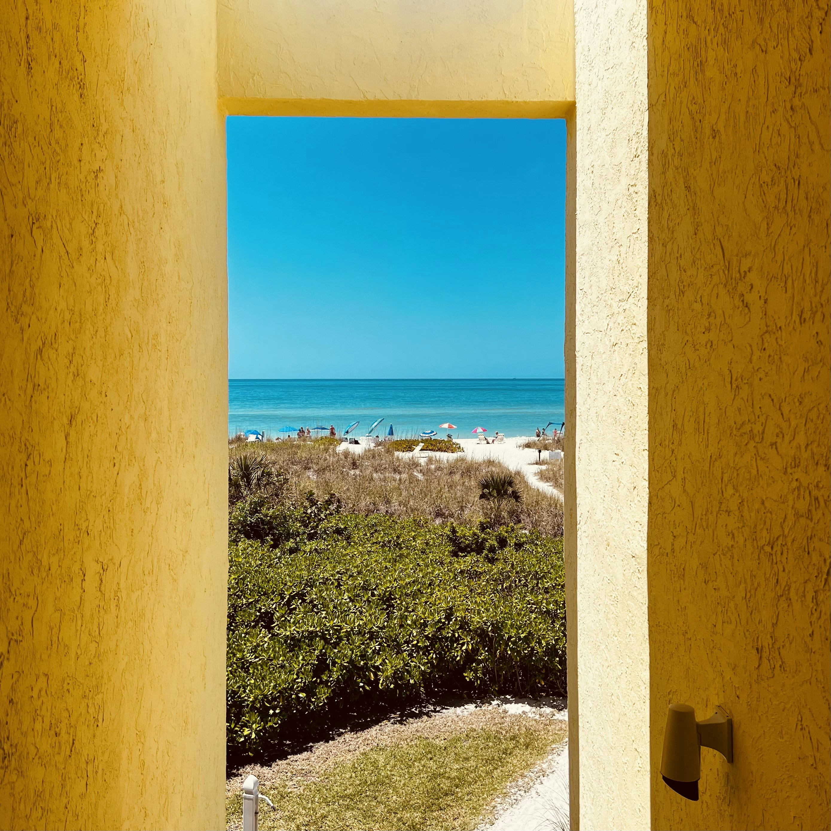 View through a yellow archway revealing a tranquil beach scene with azure waters and sunbathers. Lush greenery frames the foreground.