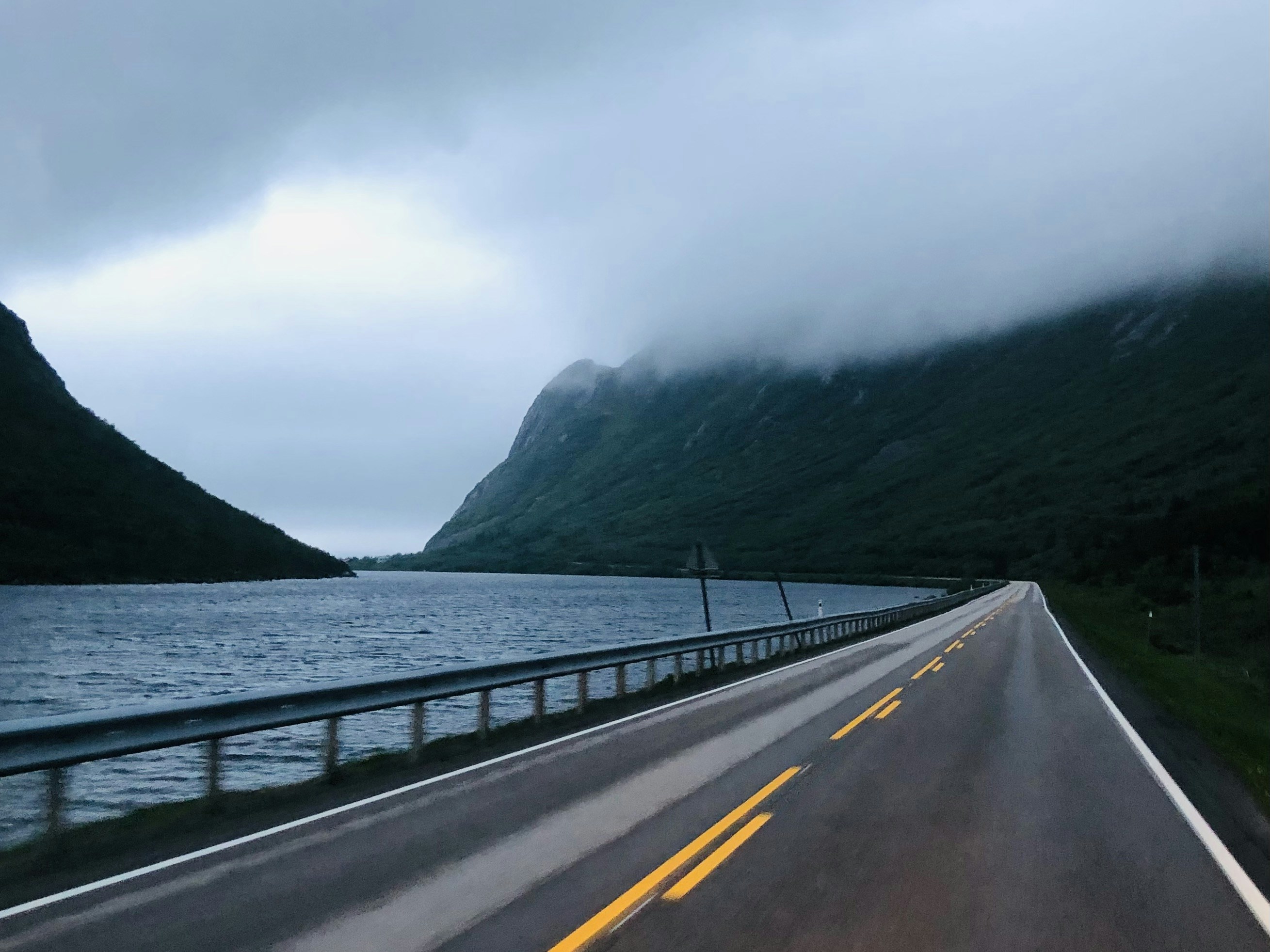 A serene road curves alongside a tranquil lake, framed by towering mountains shrouded in mist. The scene evokes a sense of adventure and exploration.