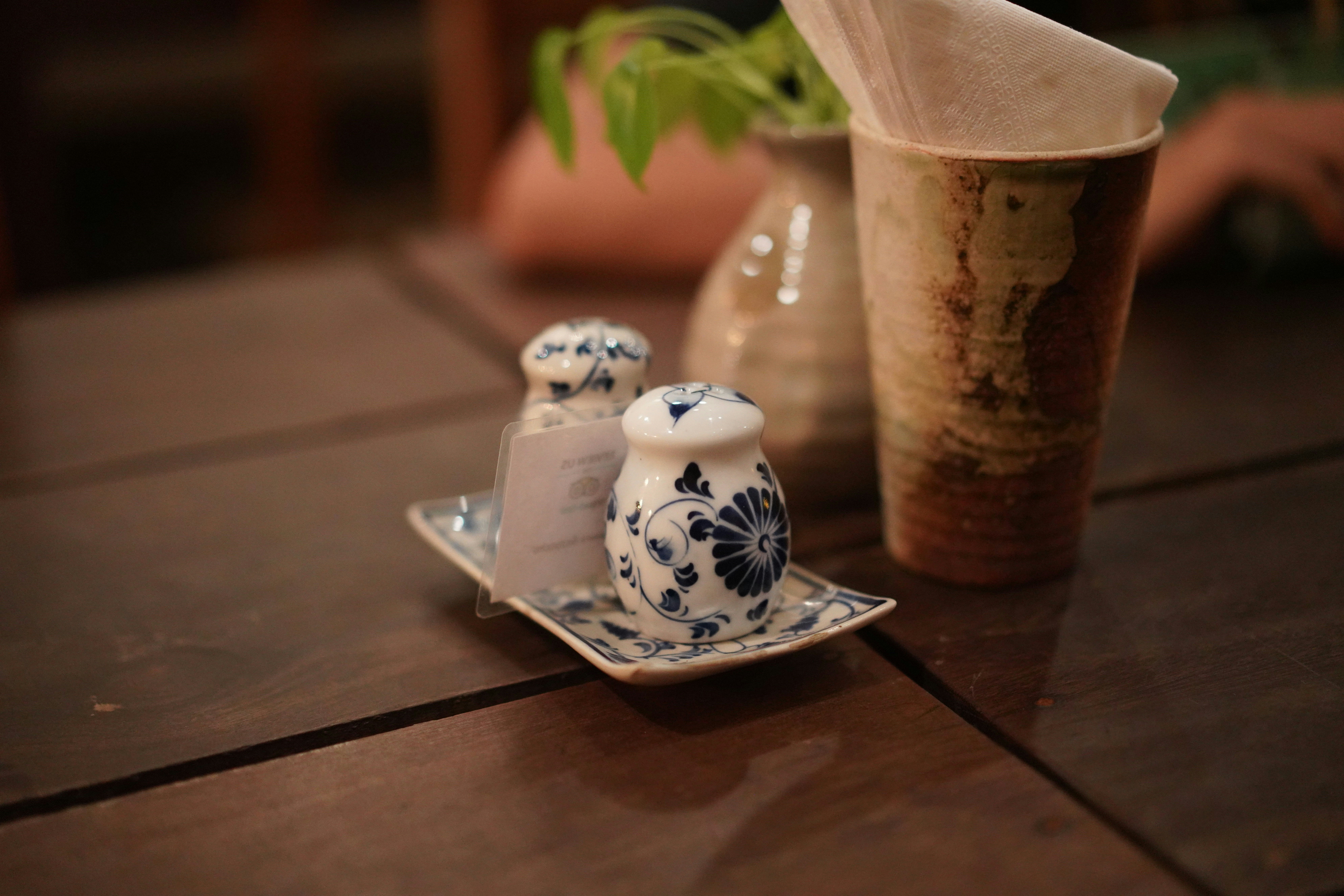 Decorative salt and pepper shakers on a plate beside a textured ceramic cup and a small plant, set against a rustic wooden table.