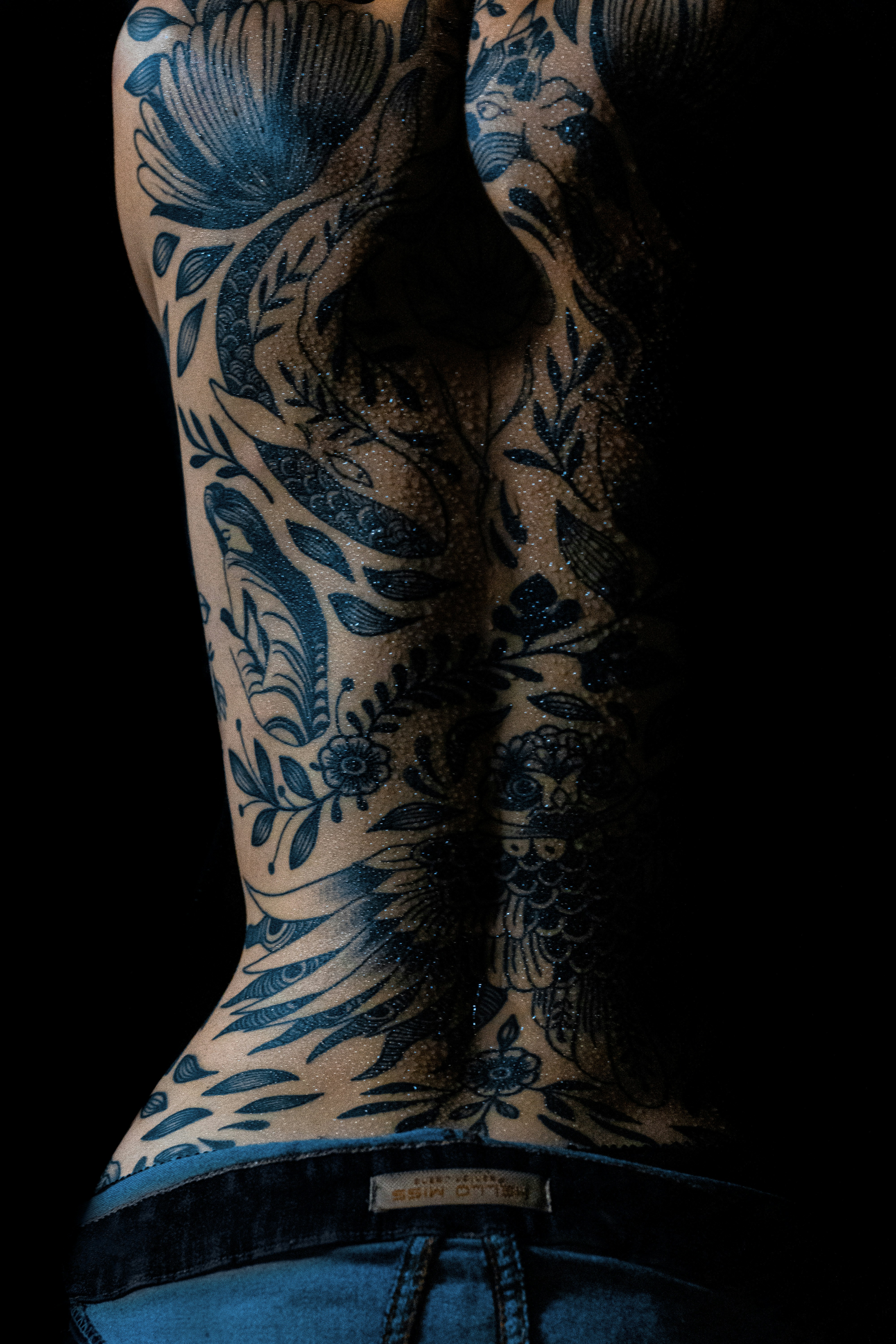 Intricate black tattoo designs adorn the back of a model, highlighting floral and animal motifs against a dark backdrop.