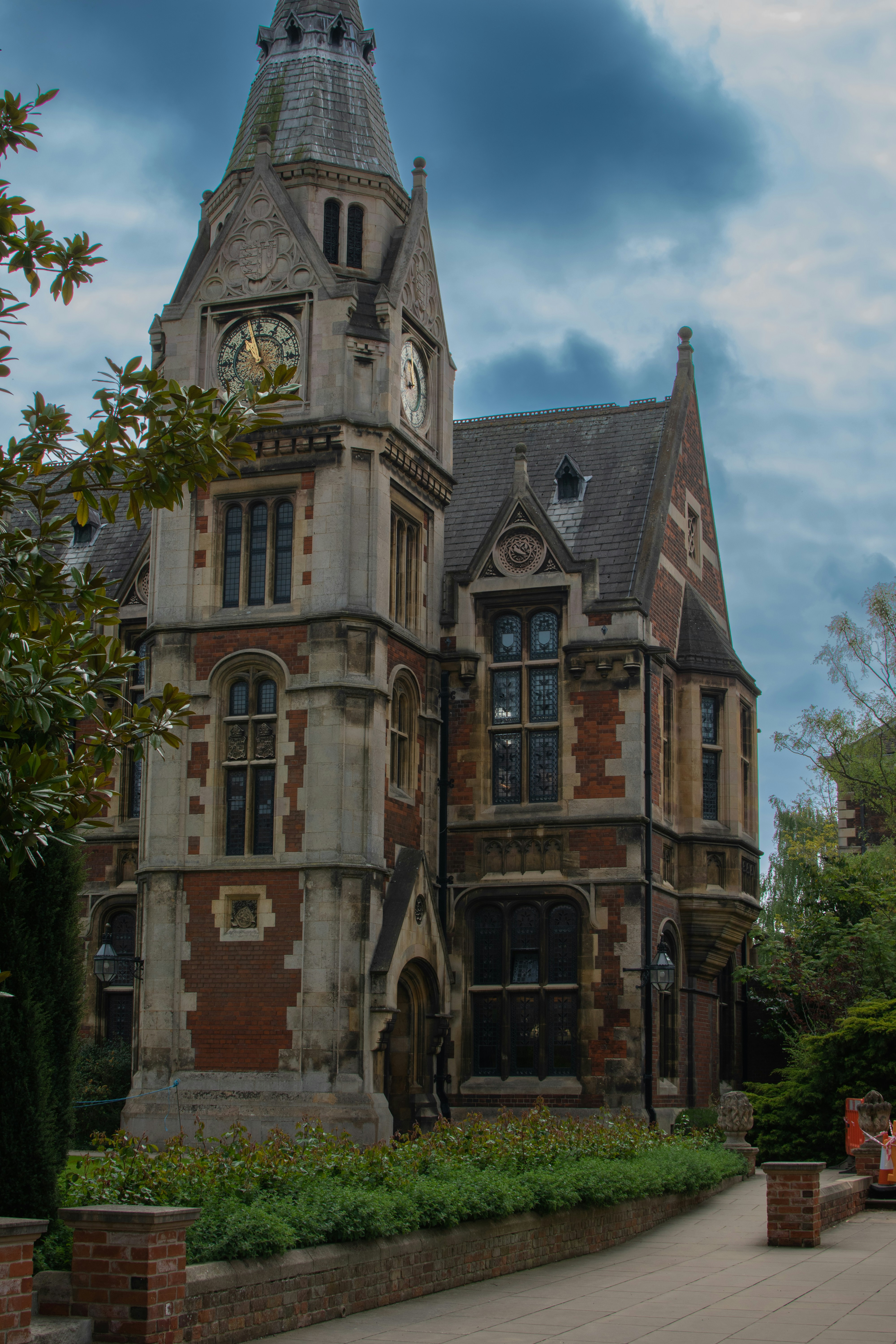 Victorian-era building featuring intricate brickwork and a prominent clock tower, surrounded by lush greenery and a serene pathway.