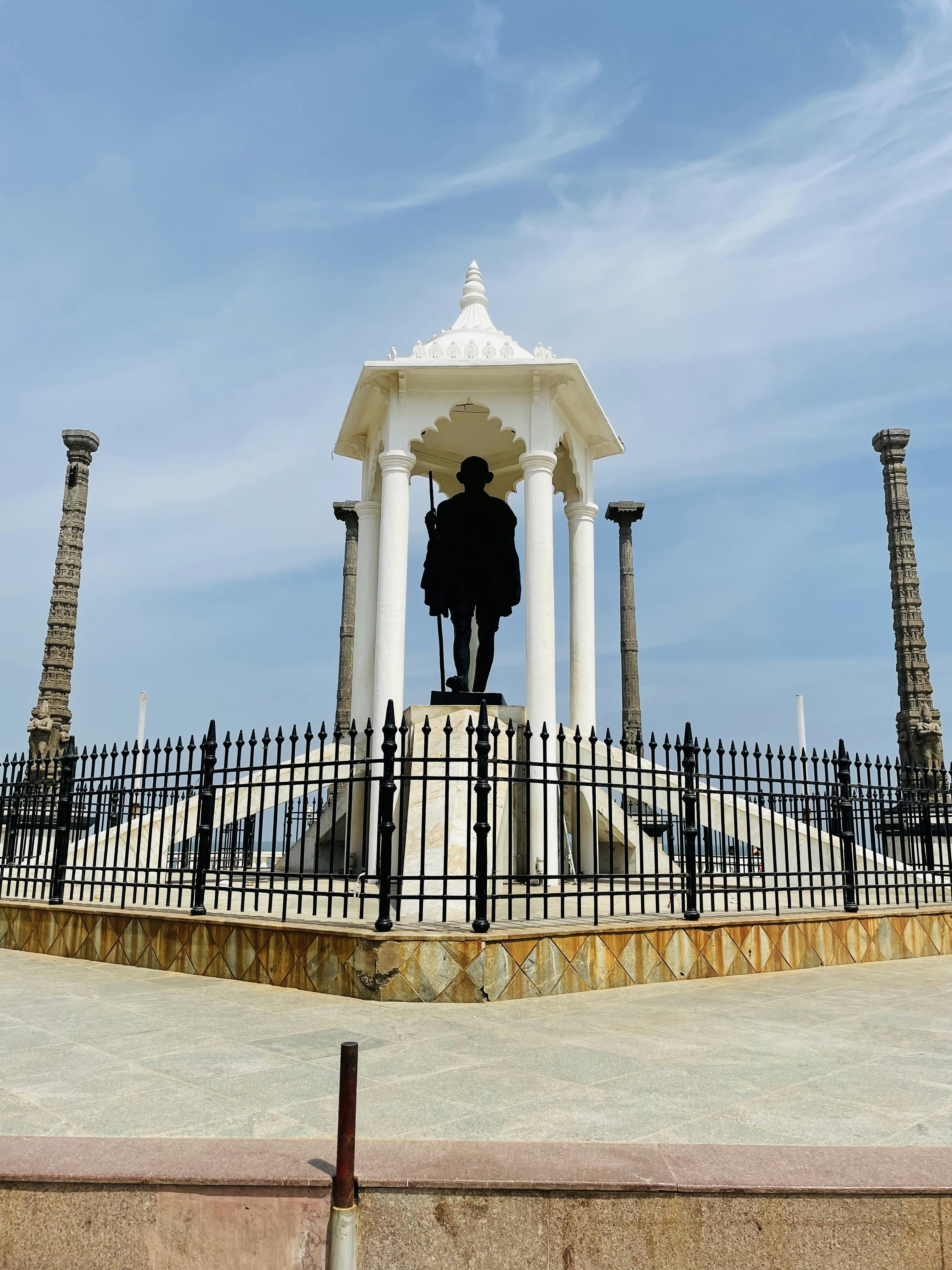 A silhouetted statue stands majestically under a white pavilion, surrounded by ornate pillars and a protective fence, symbolizing strength and resilience.