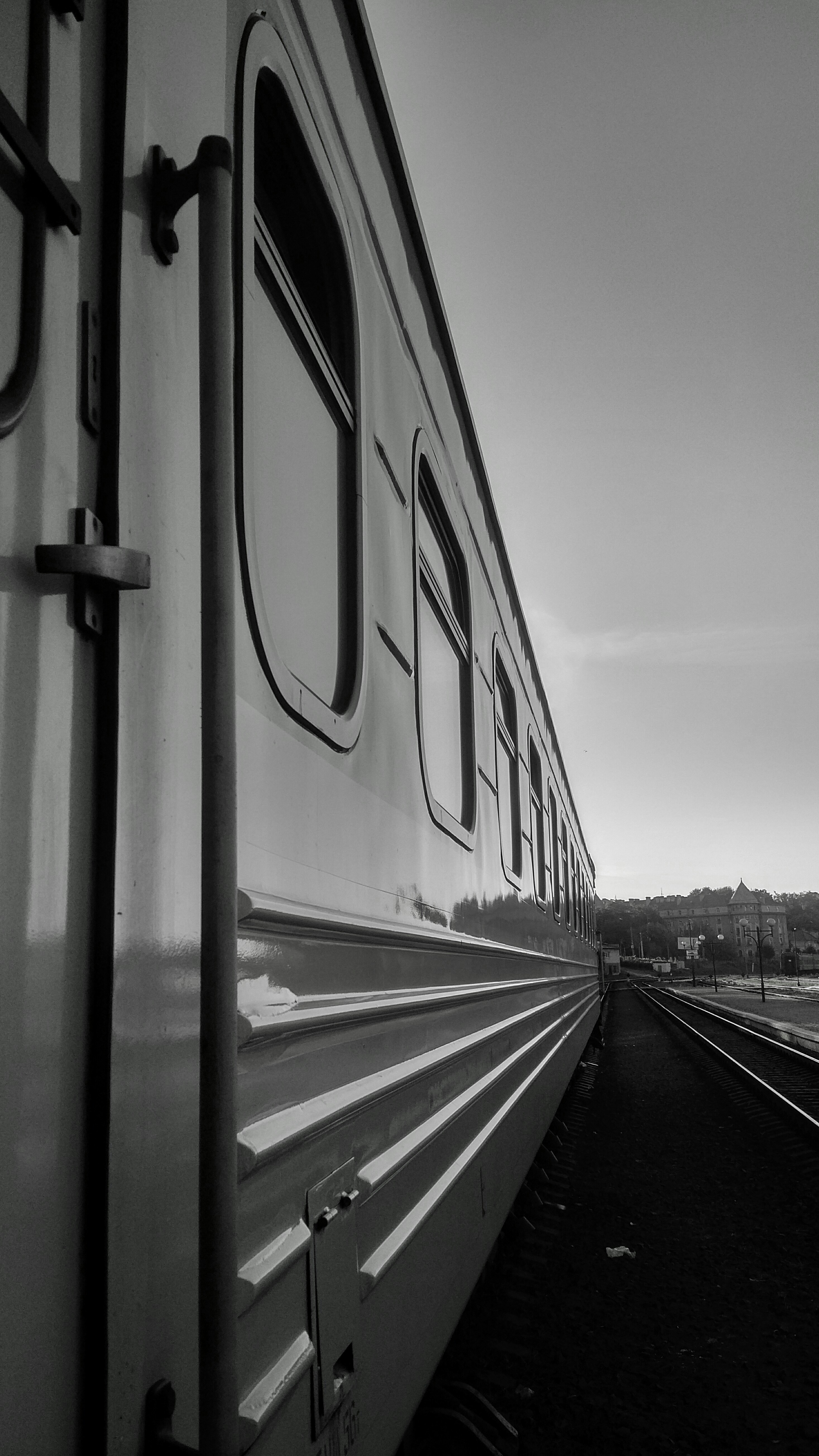 Black-and-white photograph of a train car's side, featuring repeating windows and ribbed panels that lead the eye toward the distant city along parallel tracks.