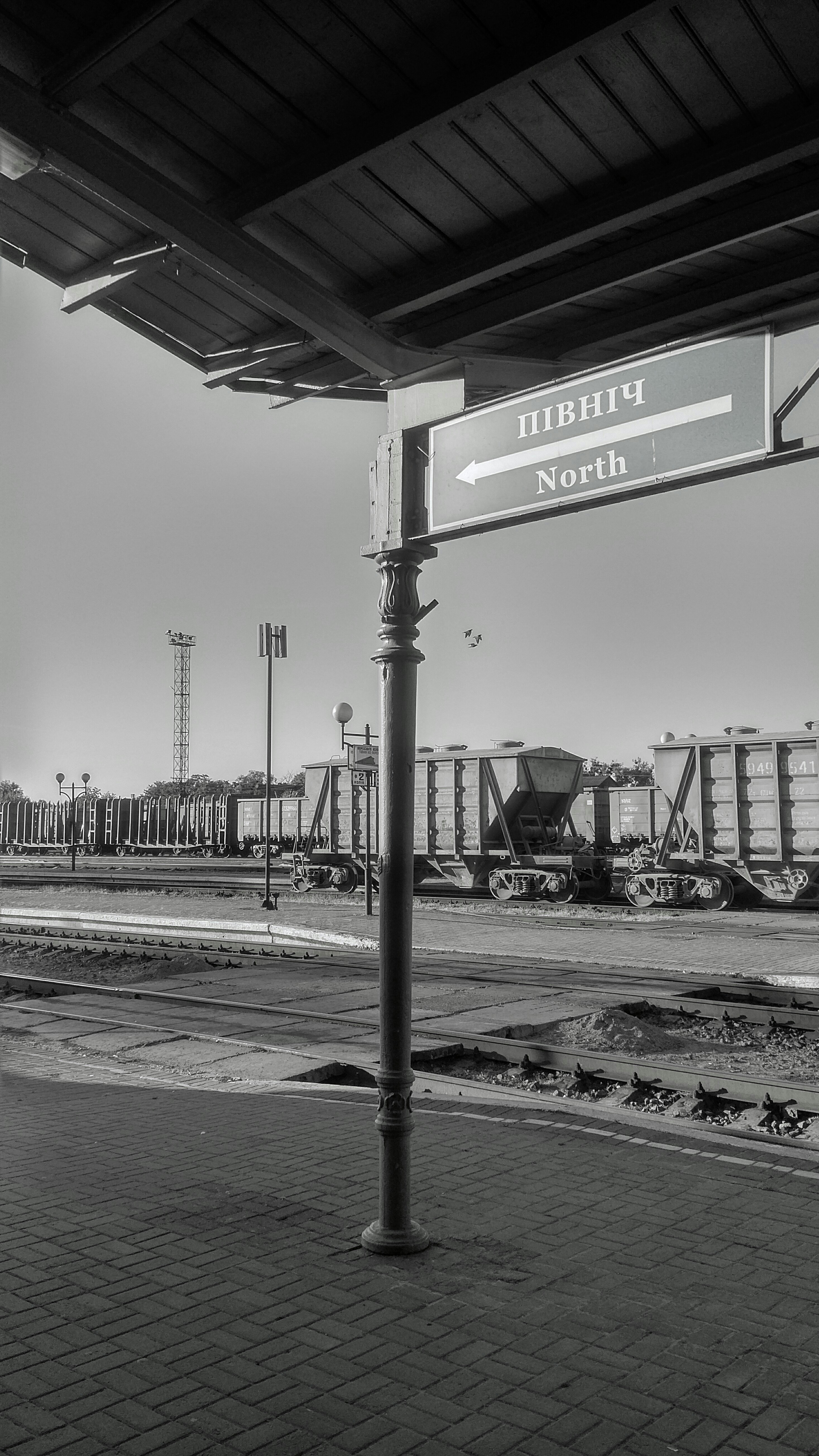 Train station signpost indicating direction to the north, with freight cars visible in the background. The scene is captured in monochrome, emphasizing the industrial atmosphere.
