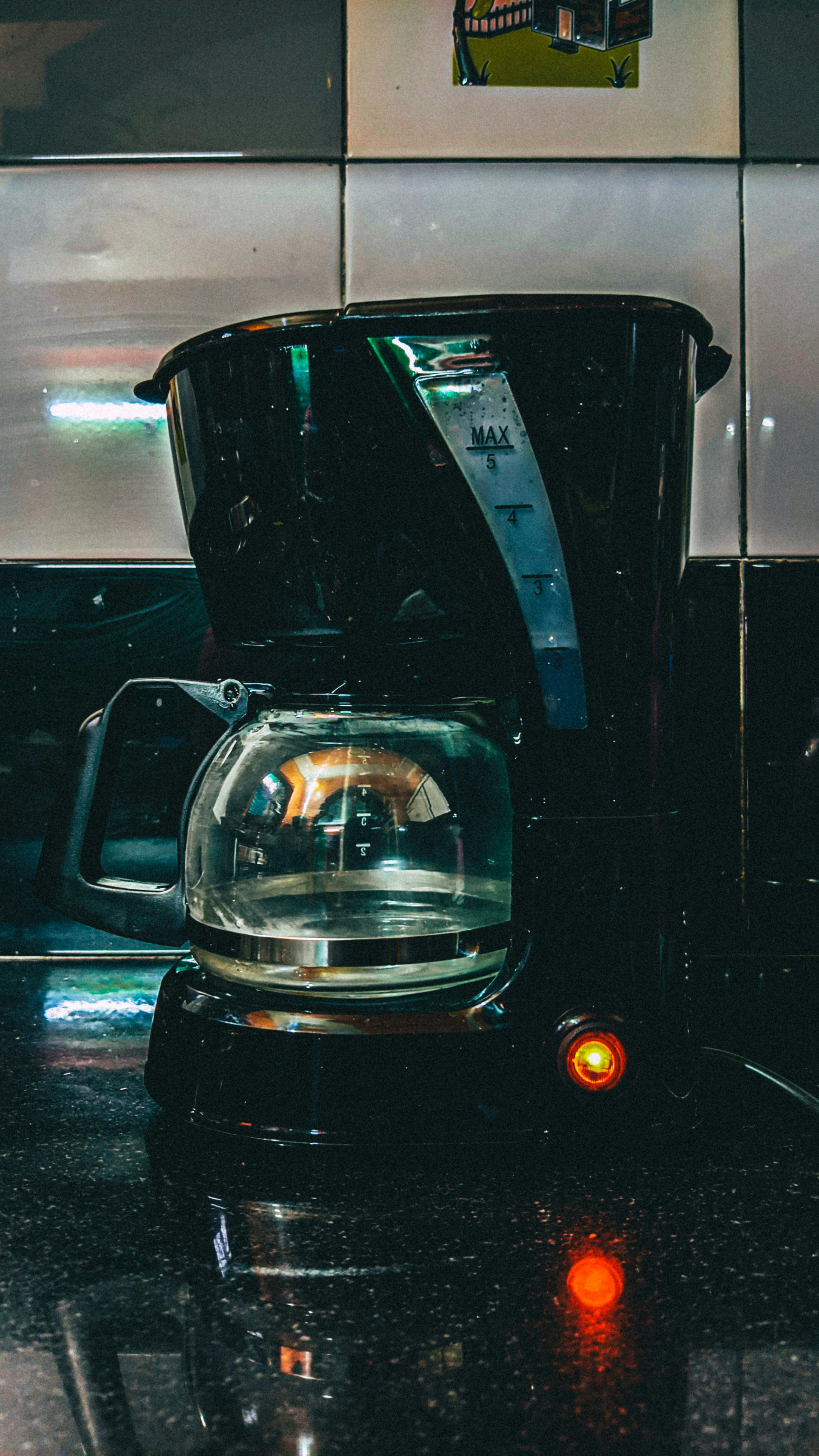 Dark-toned coffee maker with glass carafe sits on a glossy black countertop against a tiled backsplash, a warm red indicator light glows.