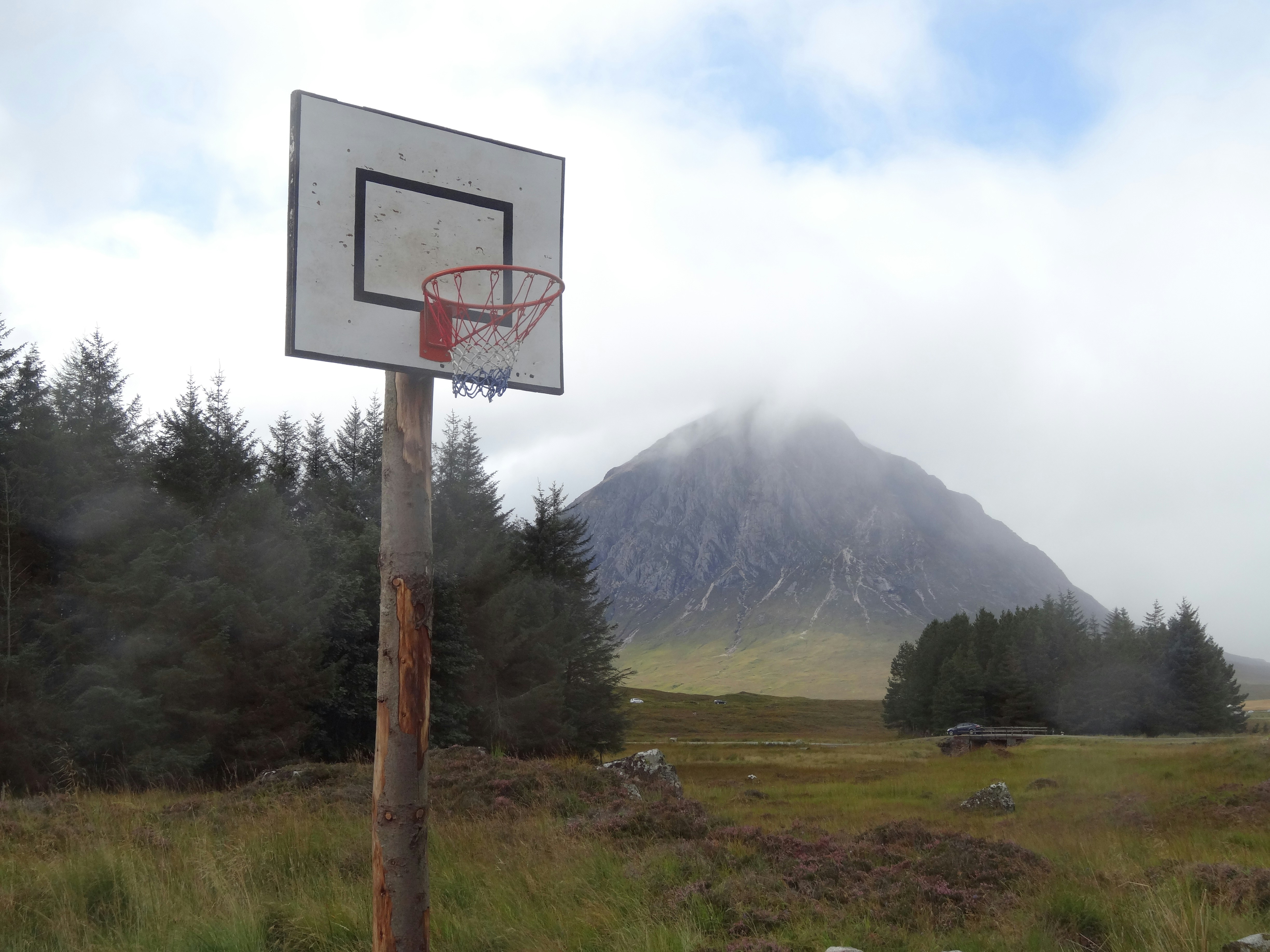 Basketball hoop stands alone in a serene landscape, framed by towering mountains and lush greenery.