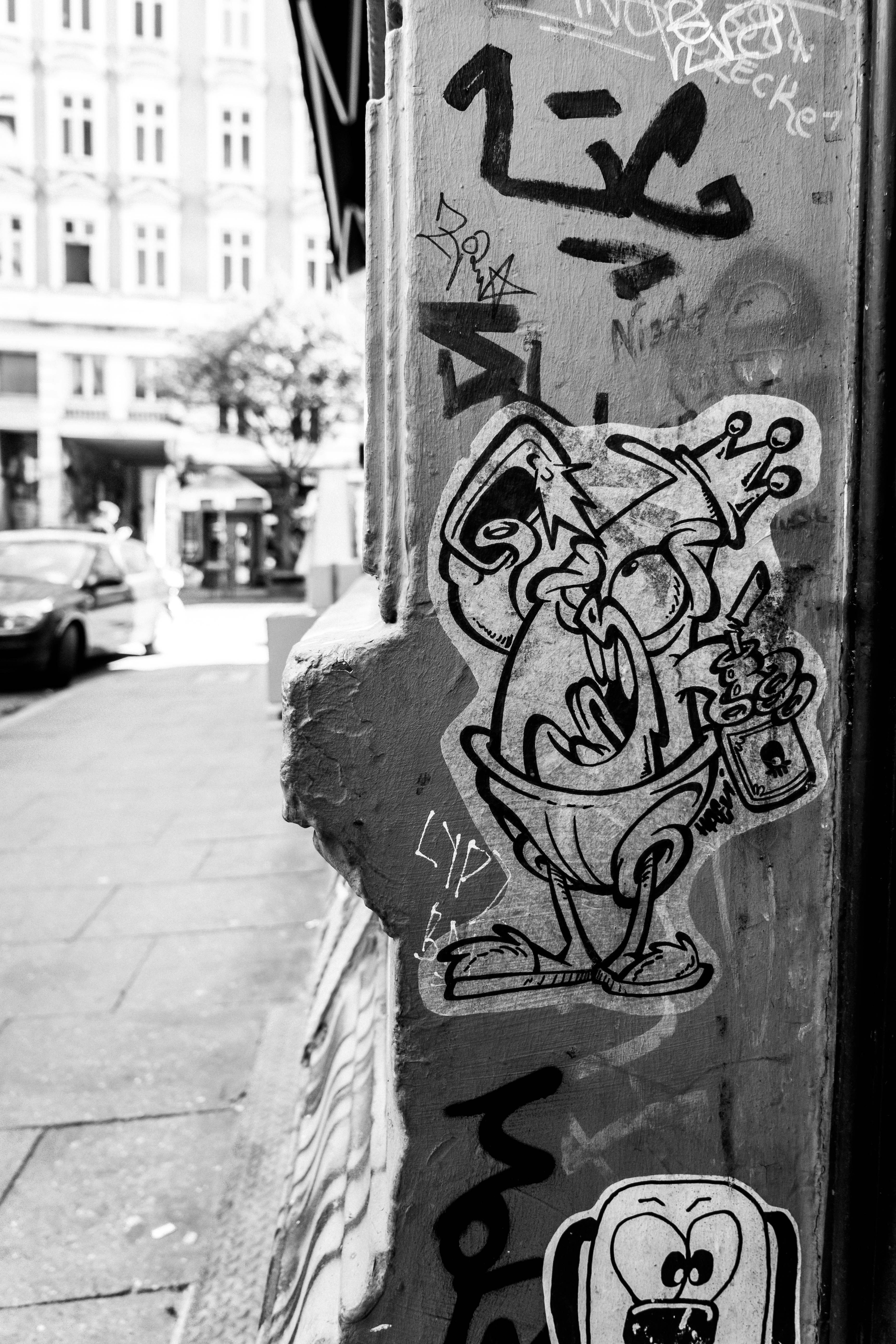 A playful cartoon character sticker adorns a weathered wall, surrounded by various graffiti tags. The scene captures the essence of urban street art culture.