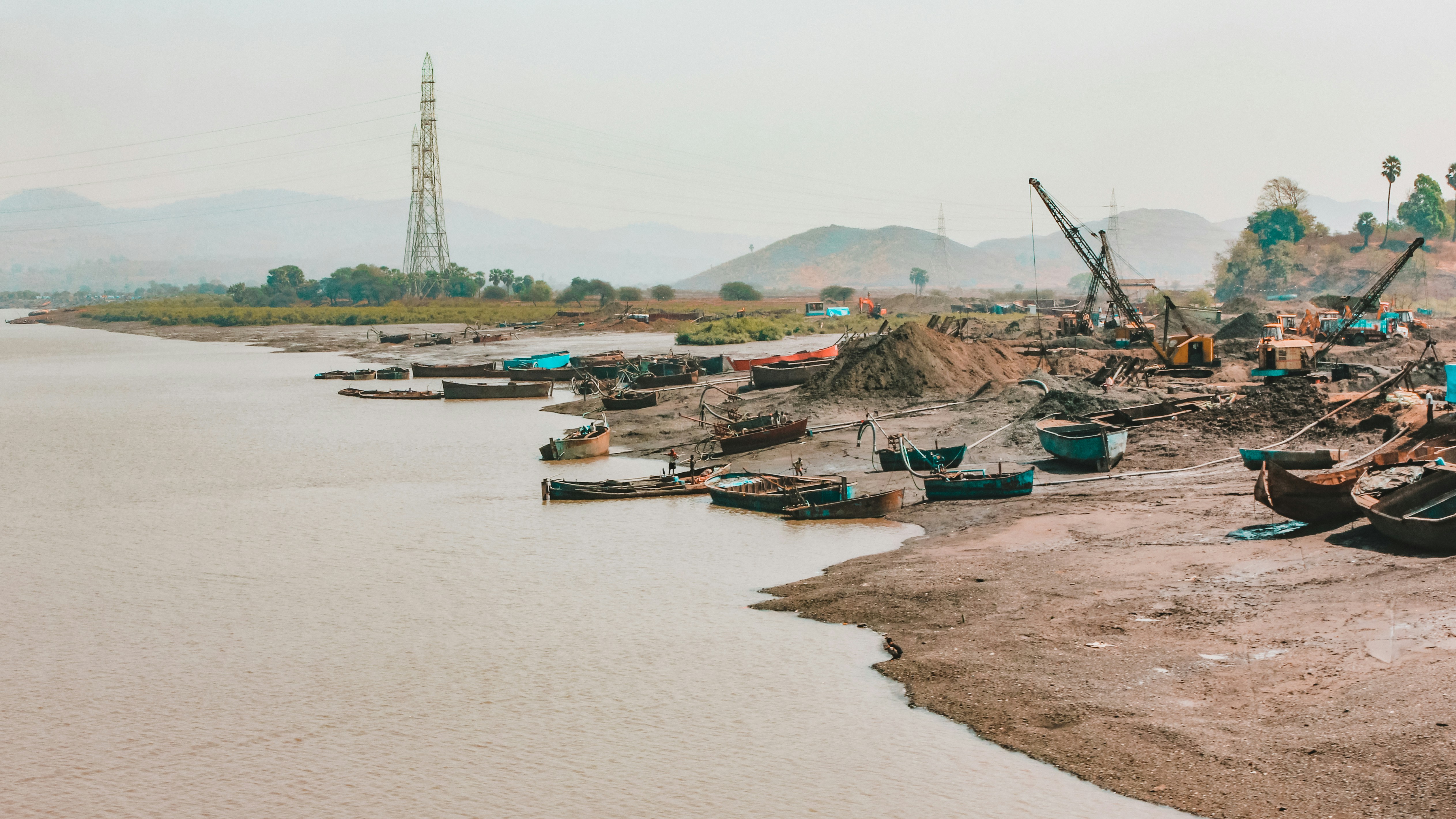 Abandoned fishing boats line a muddy riverbank, with machinery and hills in the background, illustrating the intersection of nature and human activity.