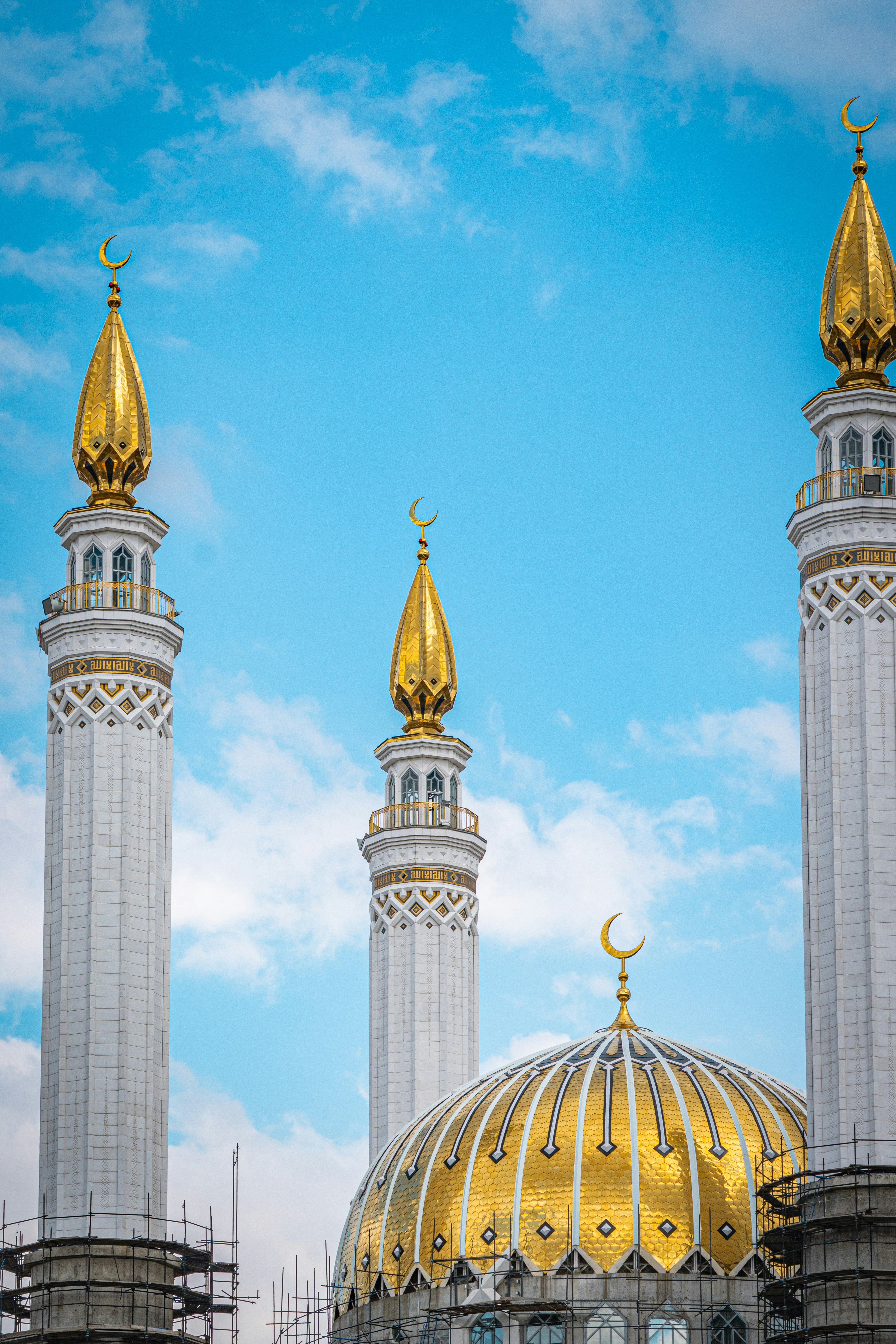 Three ornate minarets with golden crescents rise above a grand dome, framed by a clear blue sky. The architectural details reflect cultural significance.