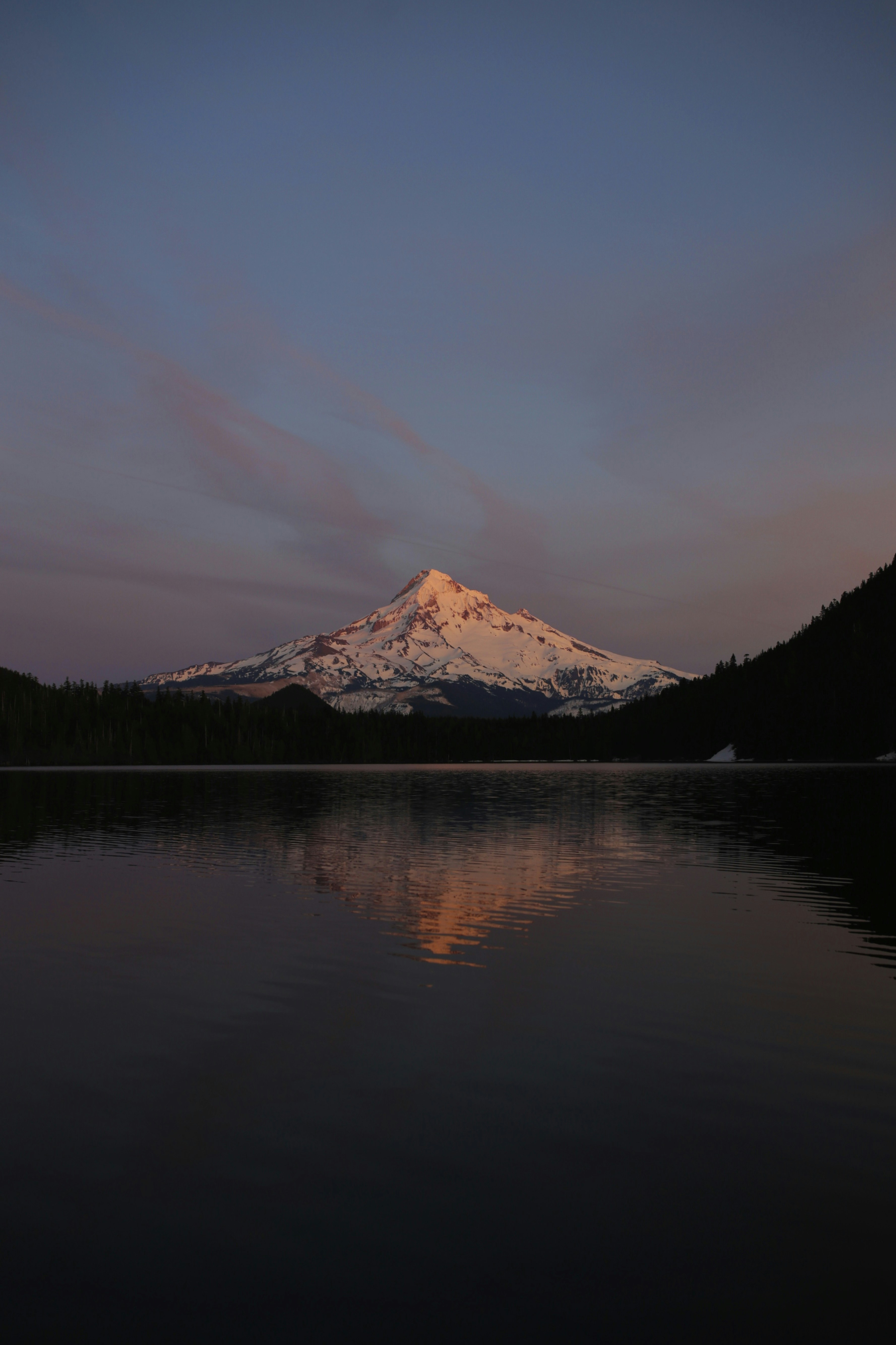 Snow-capped mountain reflecting on a tranquil lake during twilight, surrounded by lush forests.