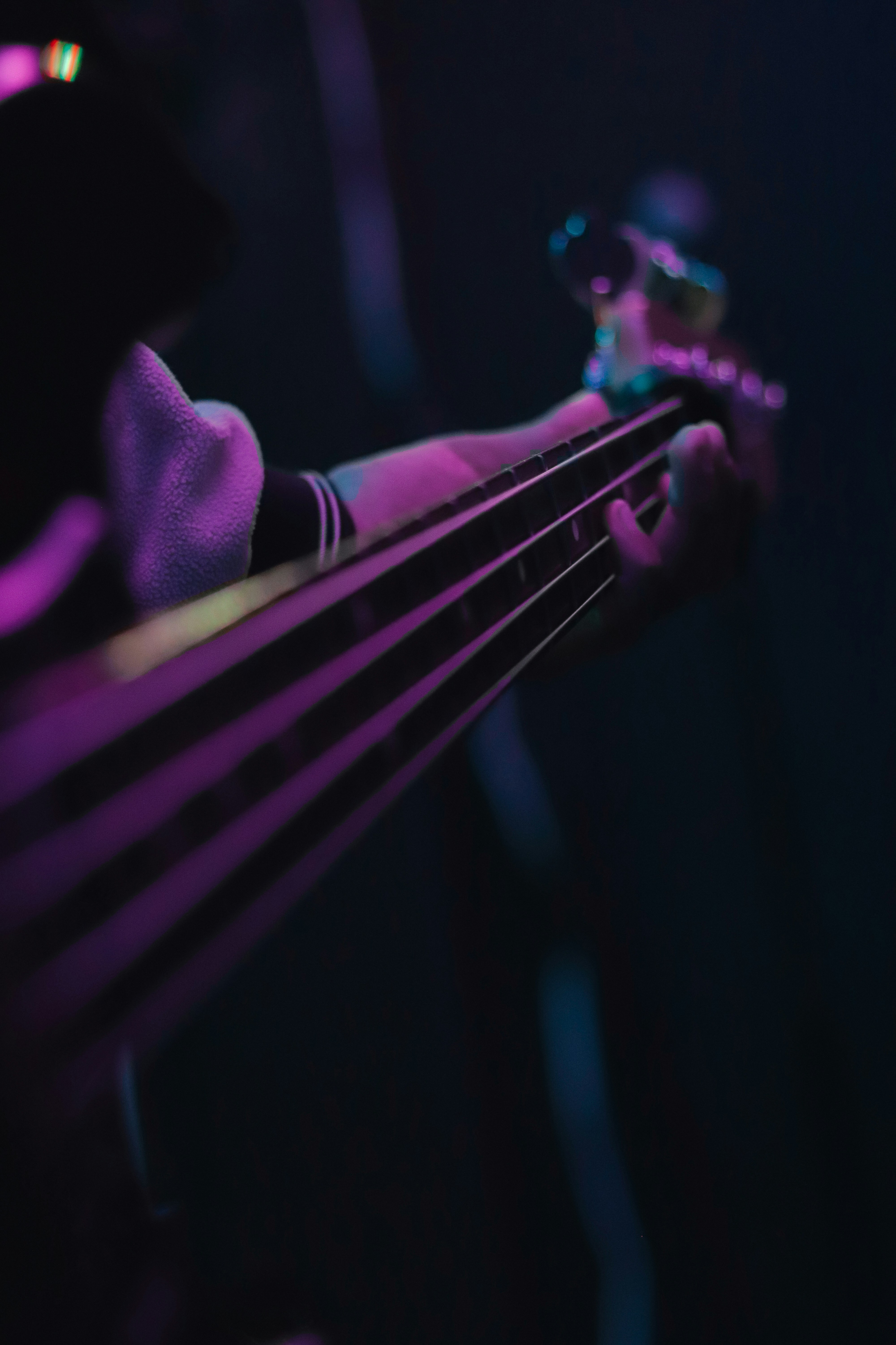 Close-up view of a hand gripping the strings of a bass guitar, illuminated by vibrant stage lights. The focus highlights the intricate details of the instrument and the musician's engagement.